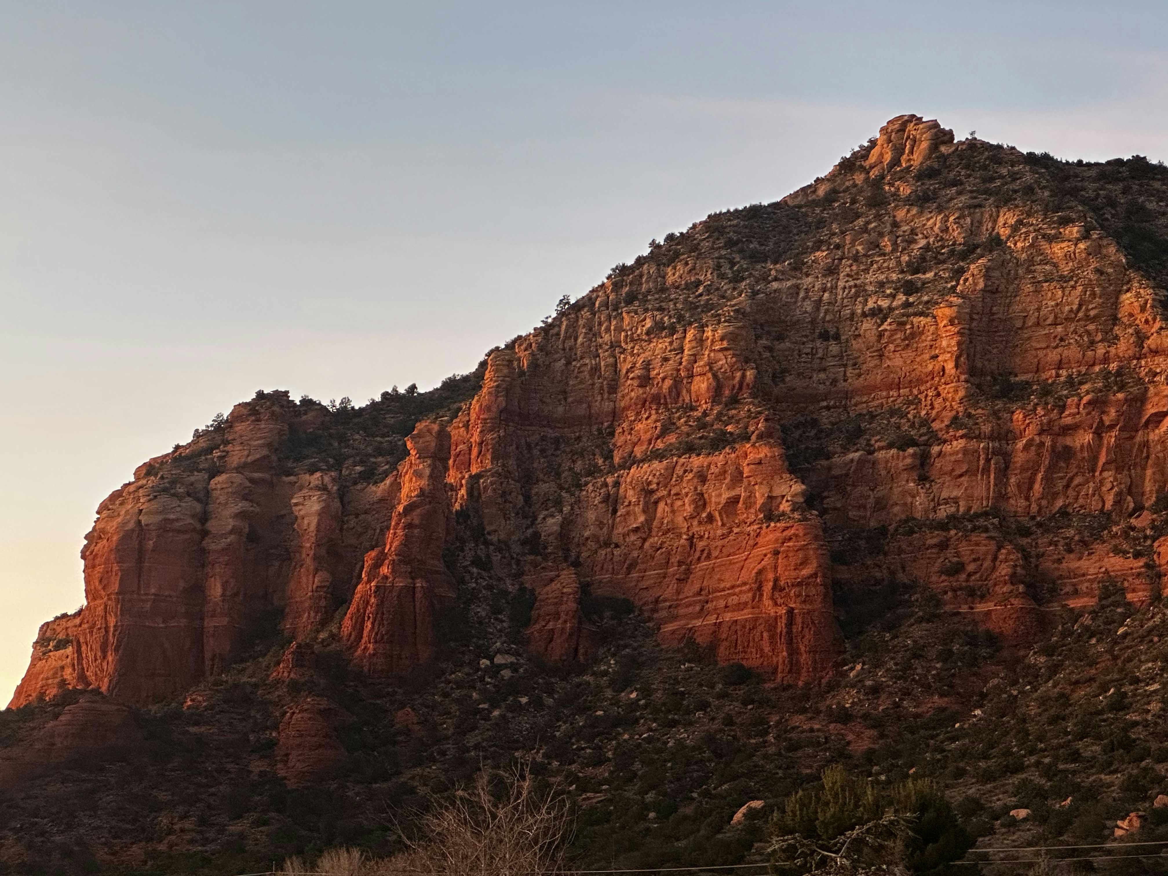 Sunlit red rock cliffs rise against a pale sky, with arid scrub at the base as evening light casts warm tones across the scene.