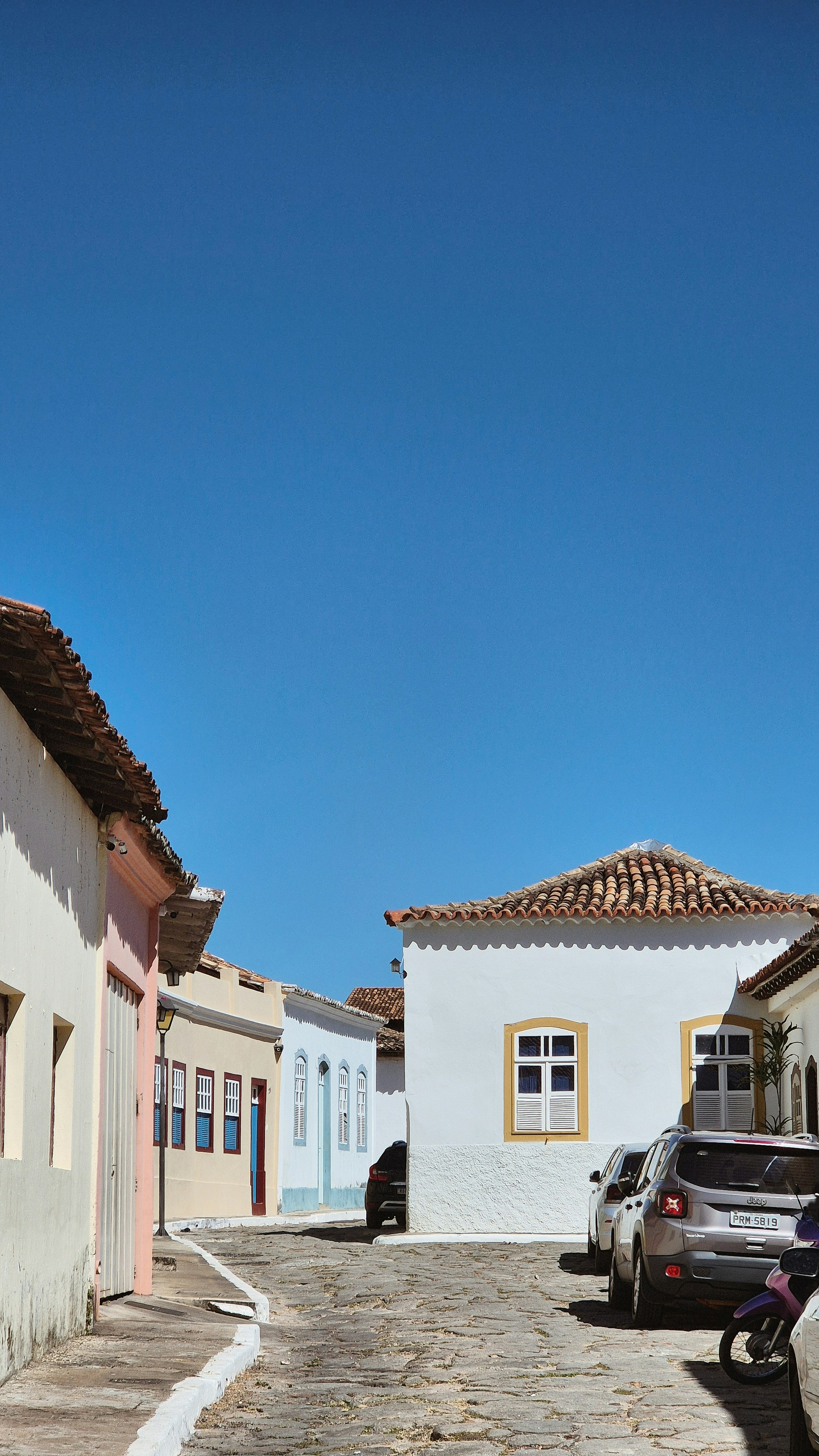 Colorful colonial houses line a cobblestone street beneath a clear blue sky, with cars parked along the right and the scene captured in natural daylight.