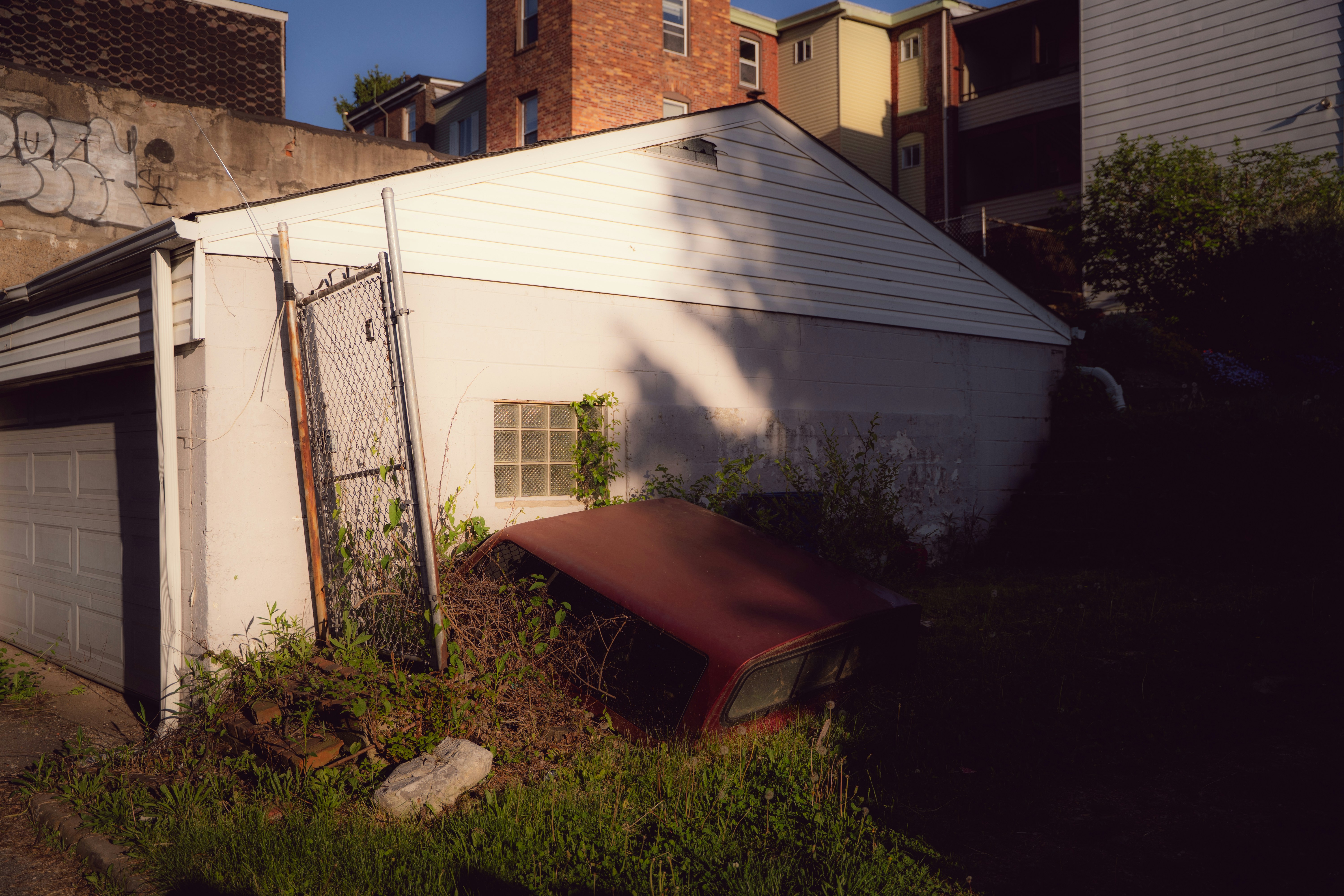 Sunlit Shadowed Garage: Abandoned Car Beneath the Leaning Fence