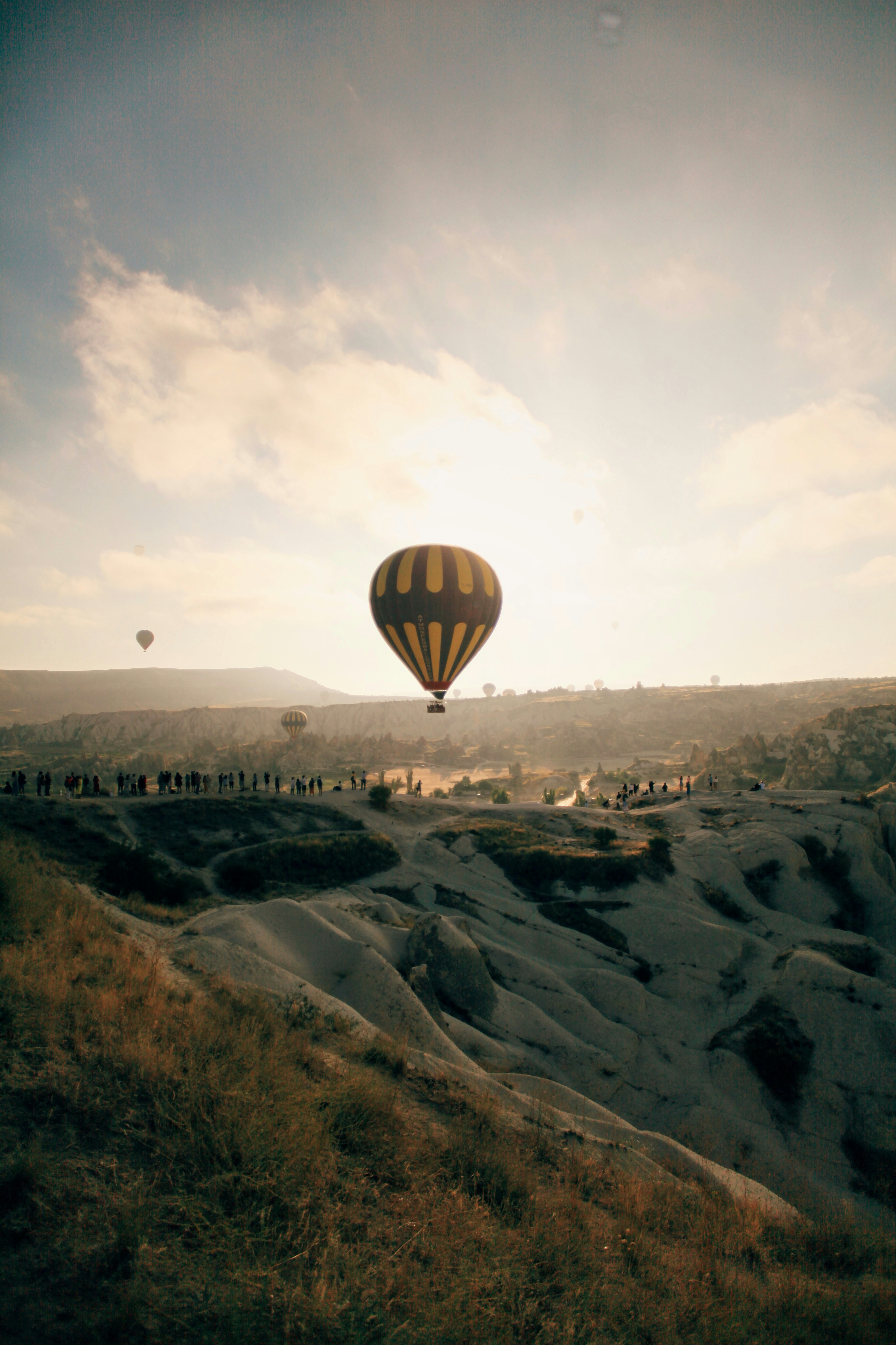 Balloons drift over a rugged canyon landscape at sunset, with a prominent hot-air balloon near center and a crowd along the ridge. A logo-like scene of travel and exploration in soft, warm tones.