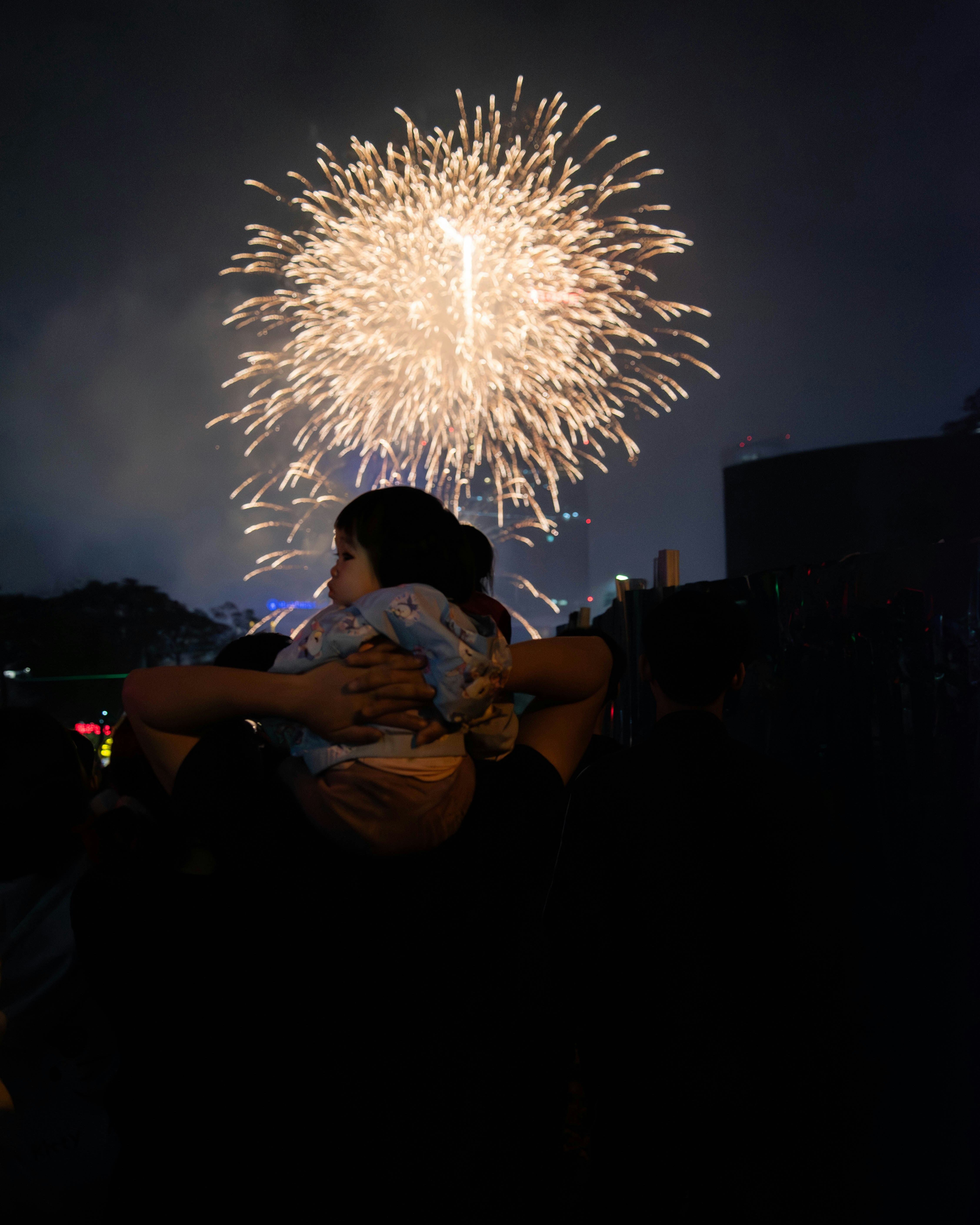 Fireworks Over Silhouetted Parade: A Child Cradled Amid Night’s Glow
