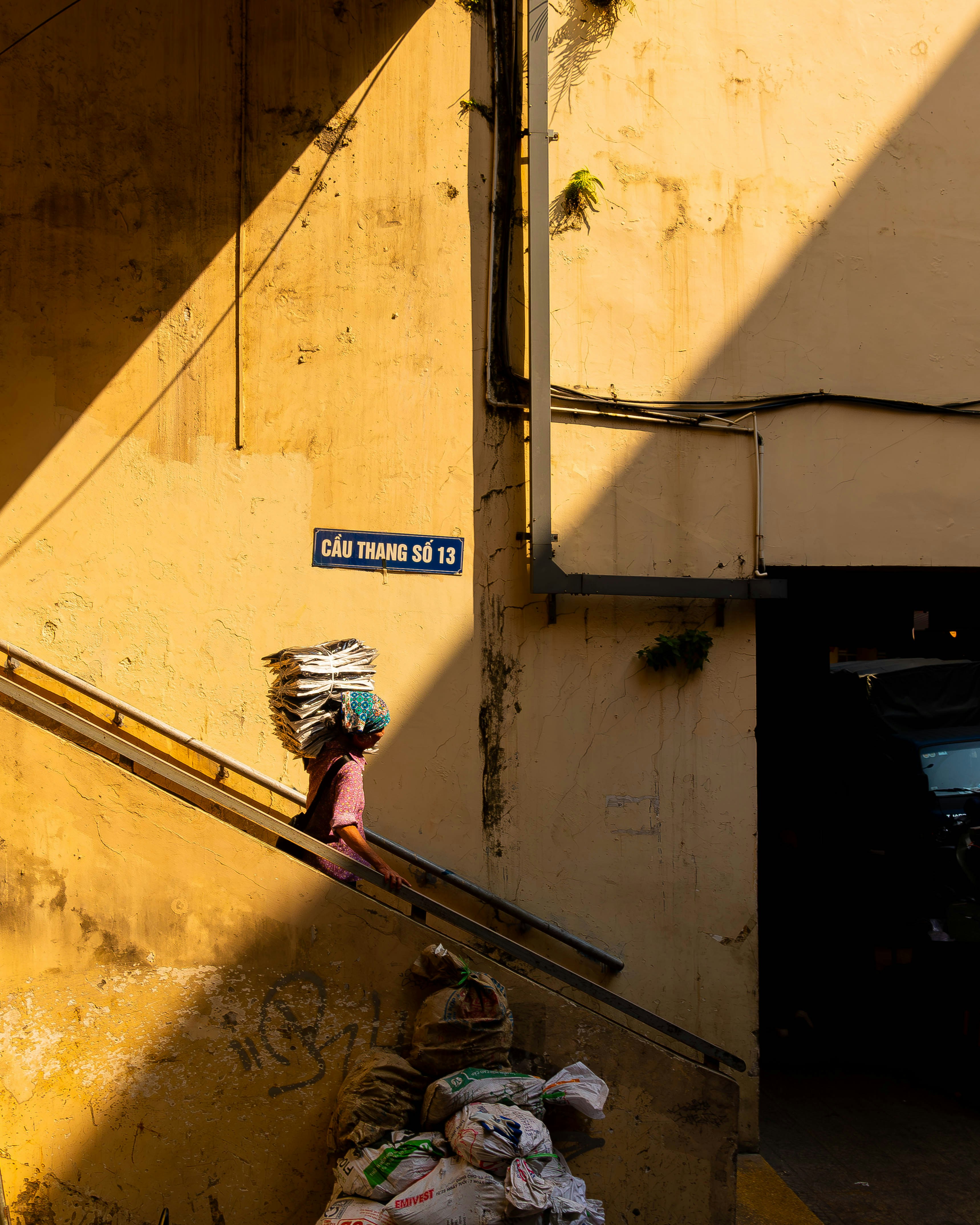 Person carrying a large bundle walks up sunlit yellow stairs.