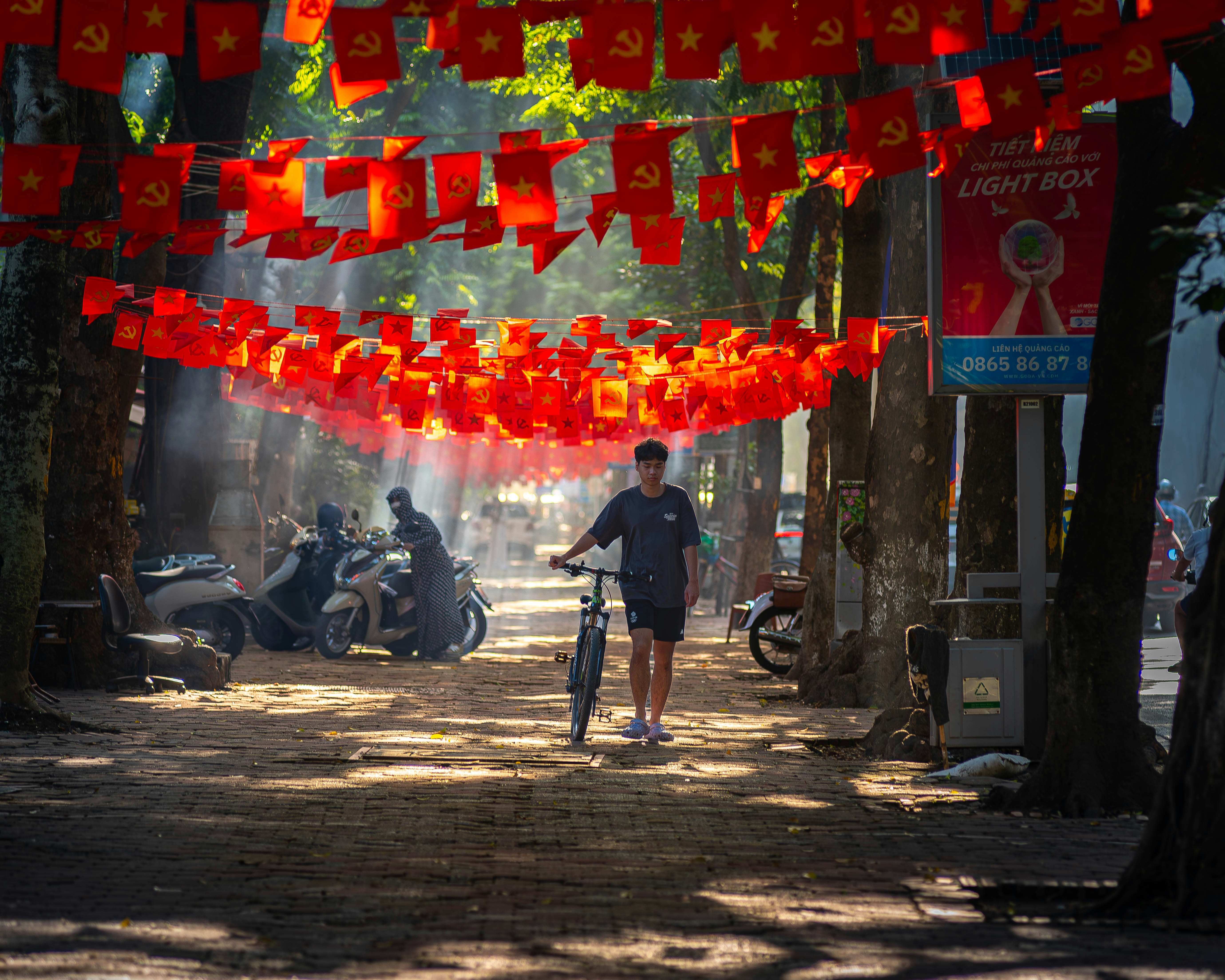 Person walking bicycle under red flags on a sunny street.