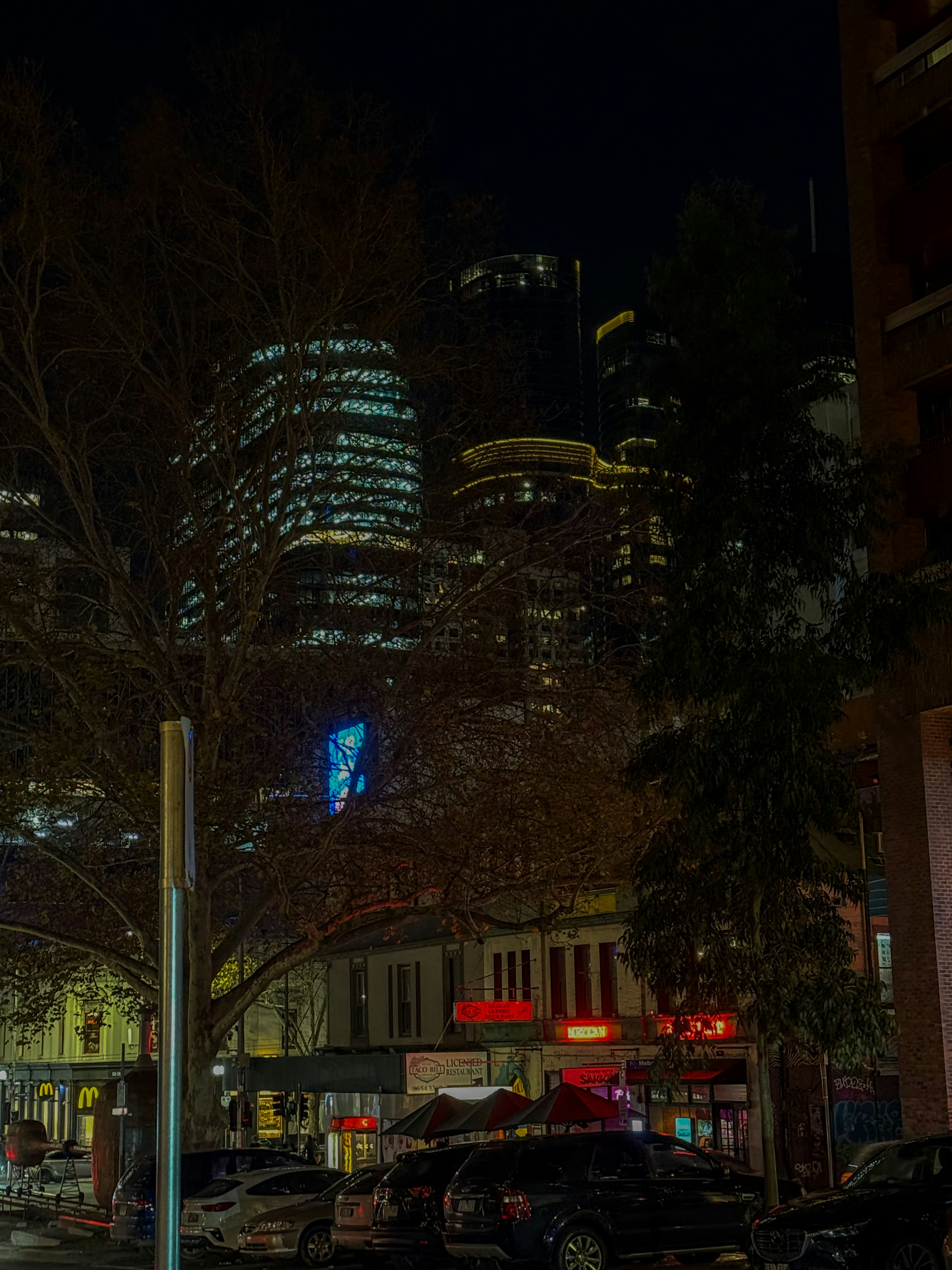 Night city street with parked cars, storefronts, and illuminated signage beneath dark trees and towering office buildings.