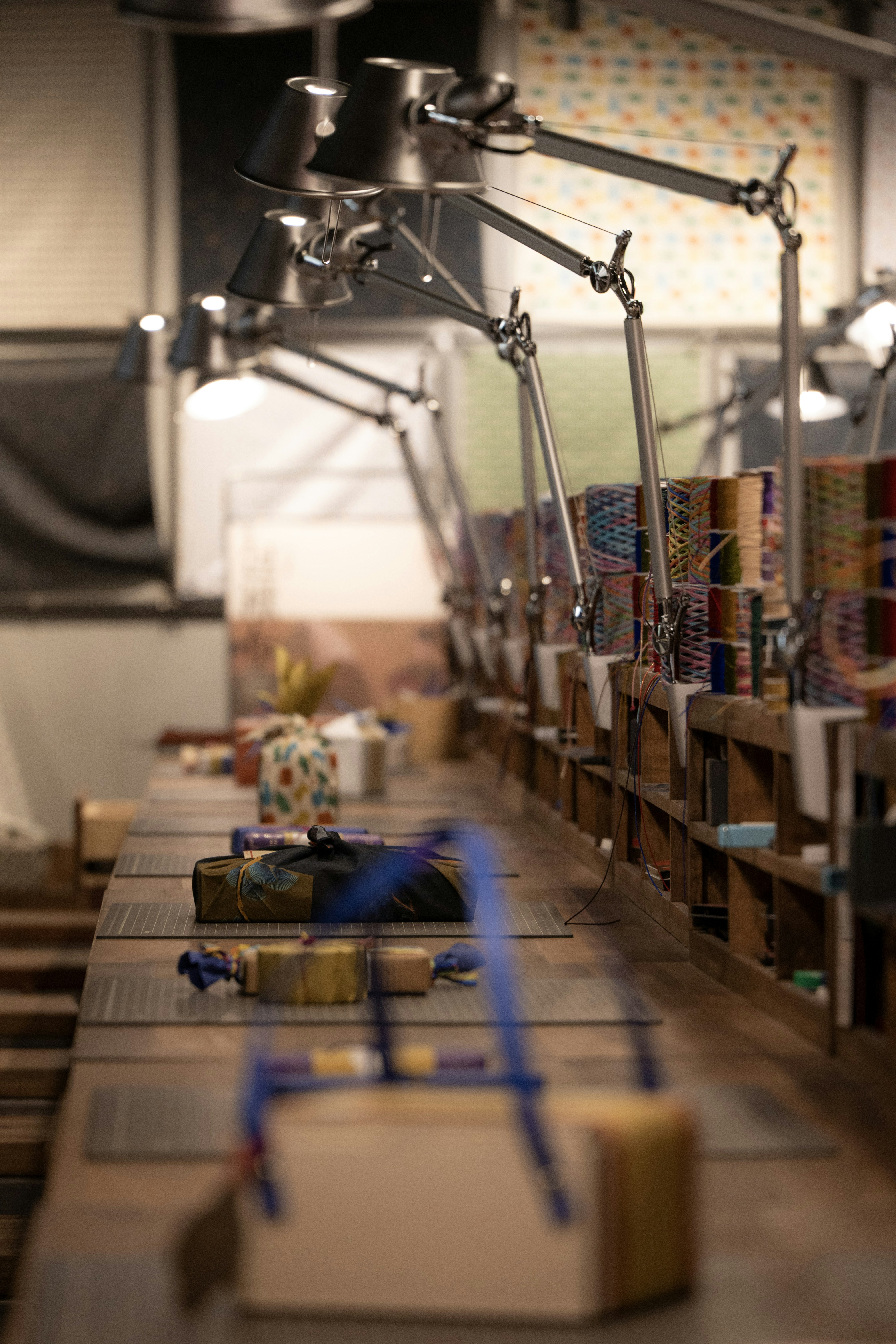 A narrow workshop aisle with a row of lamps and stacked colorful boxes along a workbench; blurs toward the foreground and background, emphasizing depth of field and the manufacturing setup.