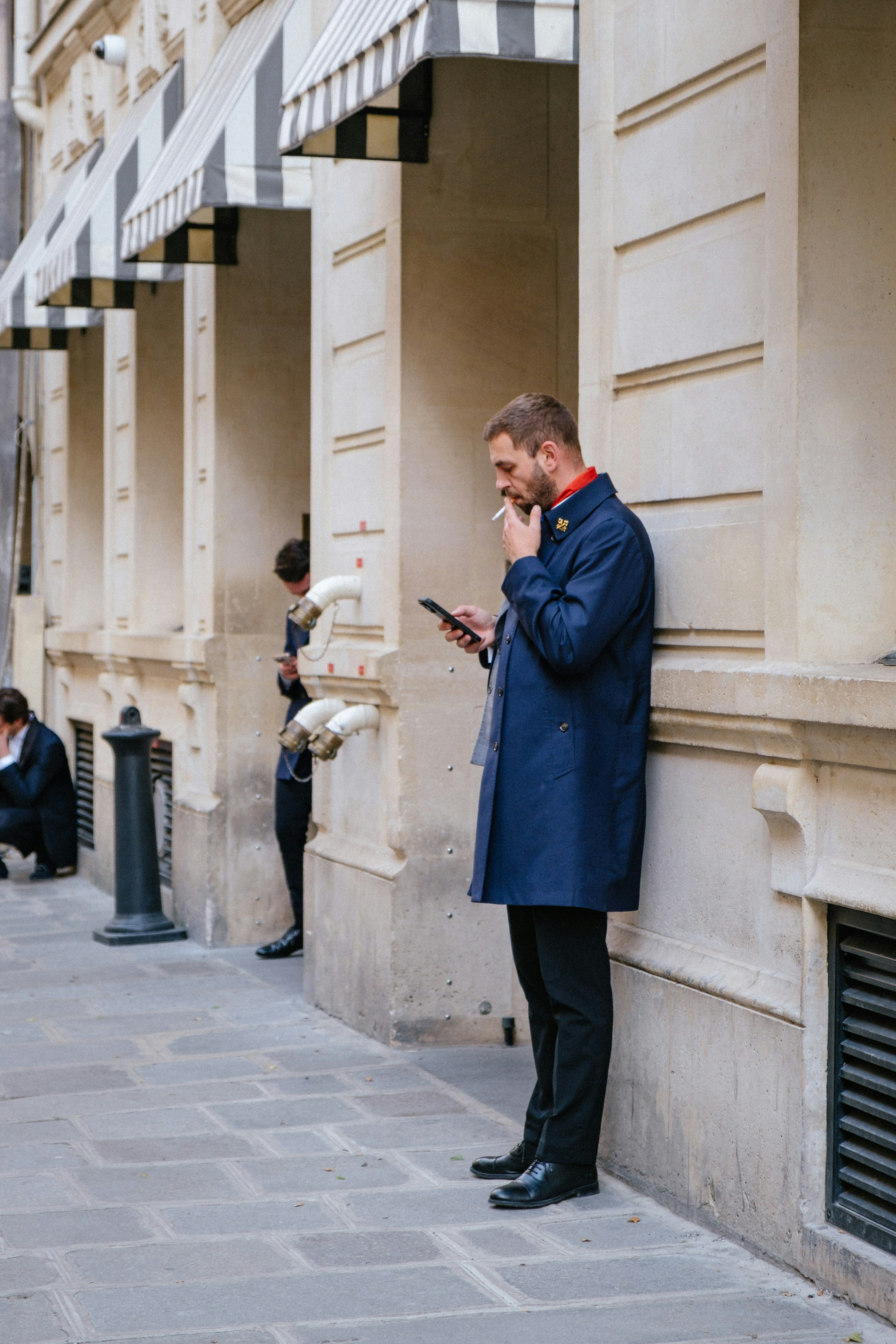 Man in a navy coat on a sunlit Parisian street checking his phone