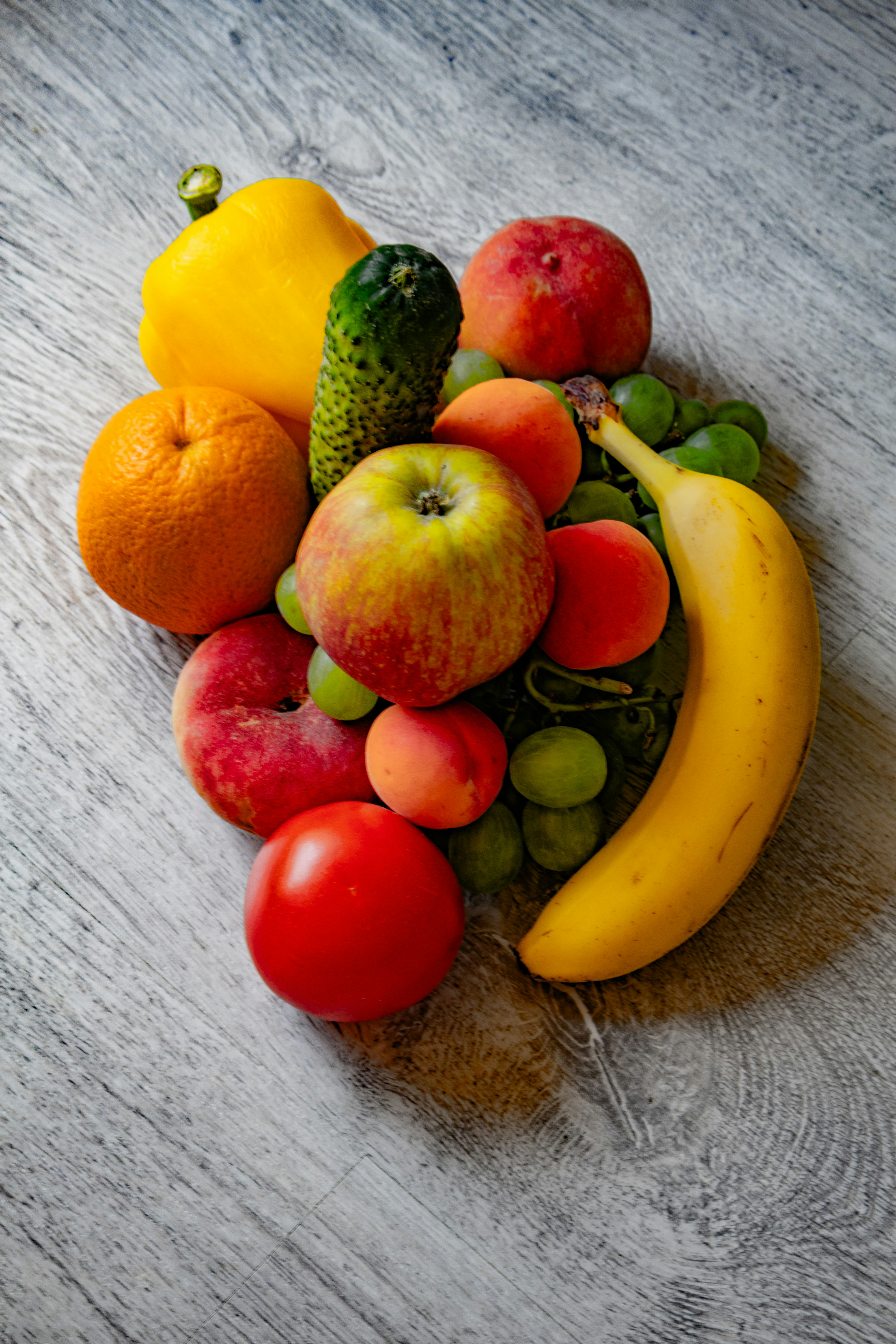 Vibrant fruit assortment arranged on a weathered surface, highlighting a banana arch and a mix of apples, oranges, grapes, and peppers; still life composition with natural textures. This is a photographic still life.