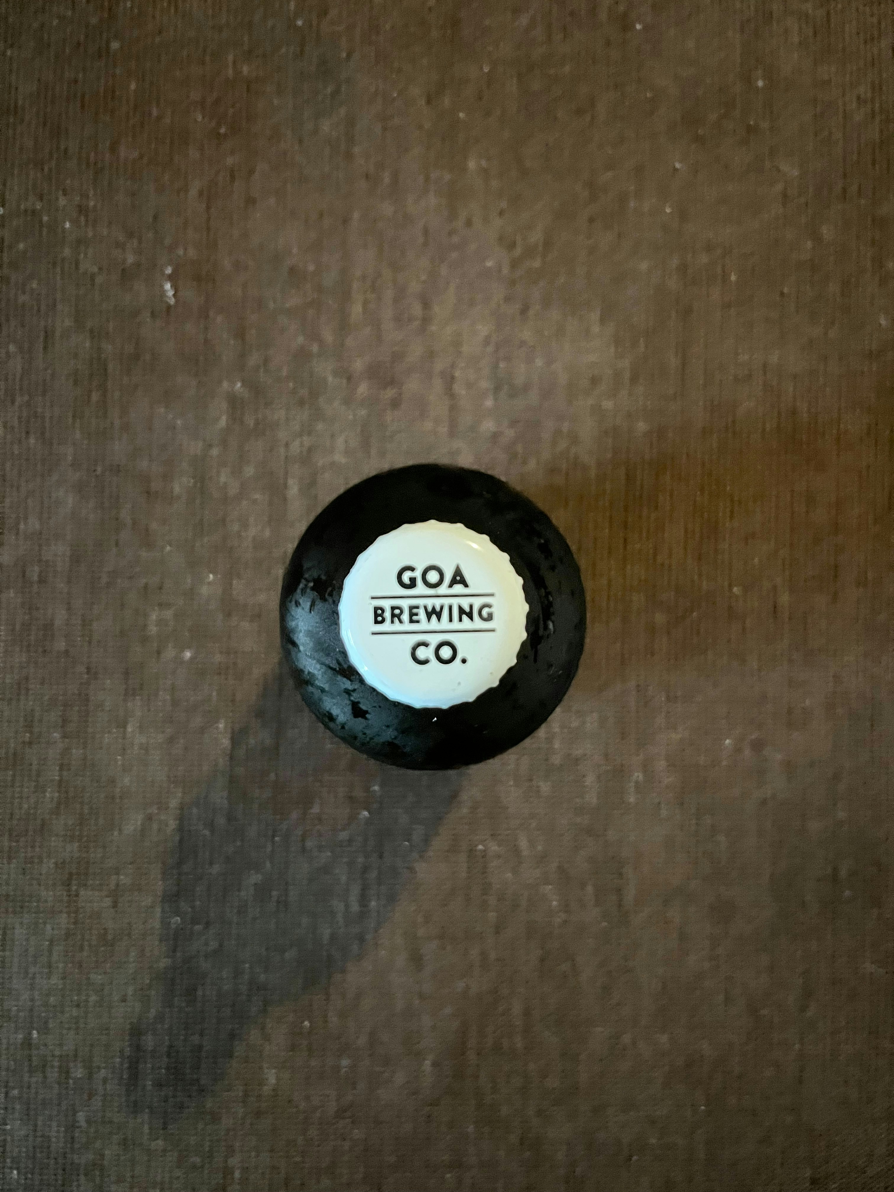 Close-up of a black bottle cap with a white Goa Brewing Co. label, resting on a dark wooden surface; the focus highlights texture and branding with subdued lighting.