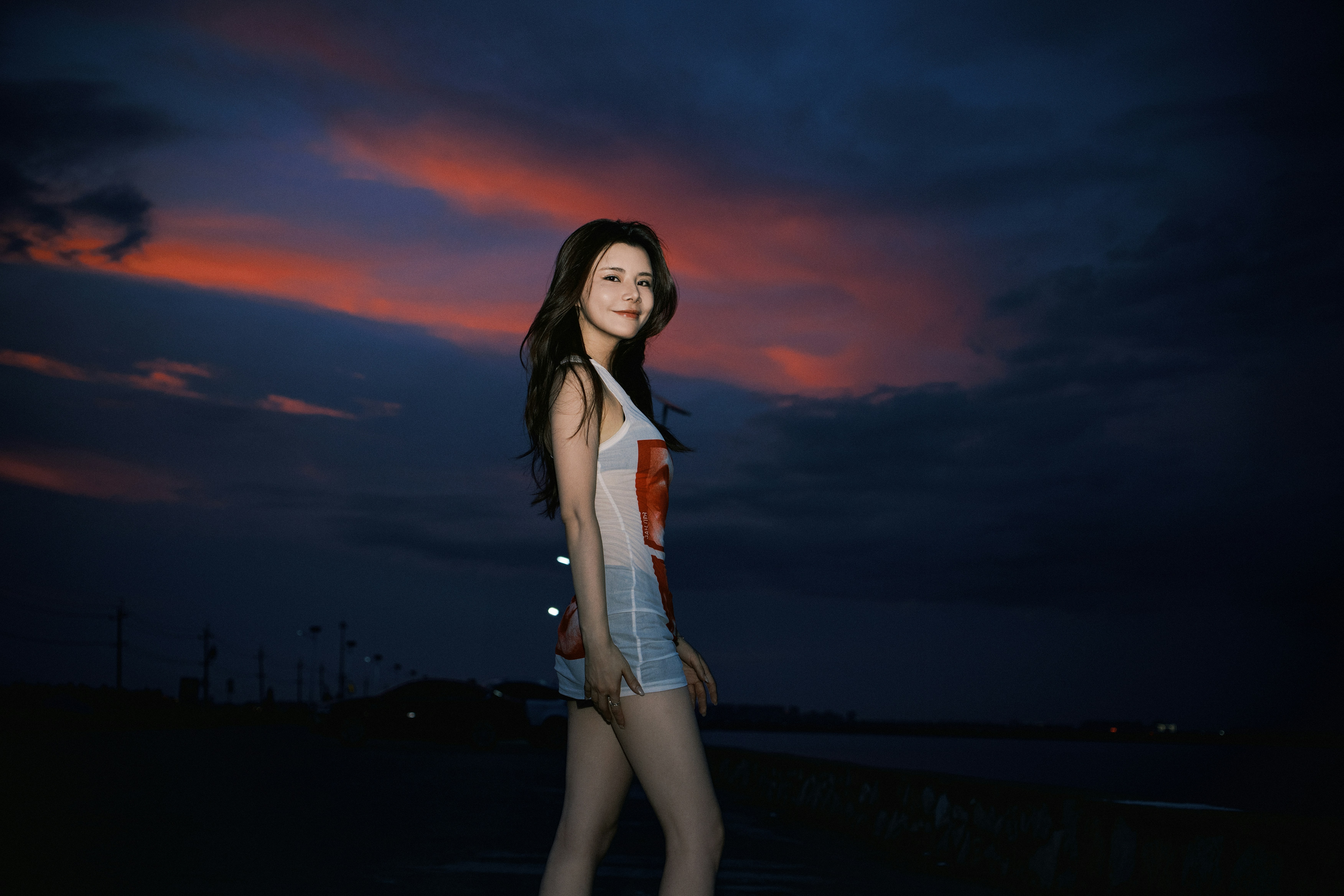 Slim figure of a young woman stands at night by the water with a dusky sunset glow behind her, wearing a sleeveless top and shorts. The image portrays a moody, low-light portrait with a distant horizon and subtle artificial lighting on the subject.