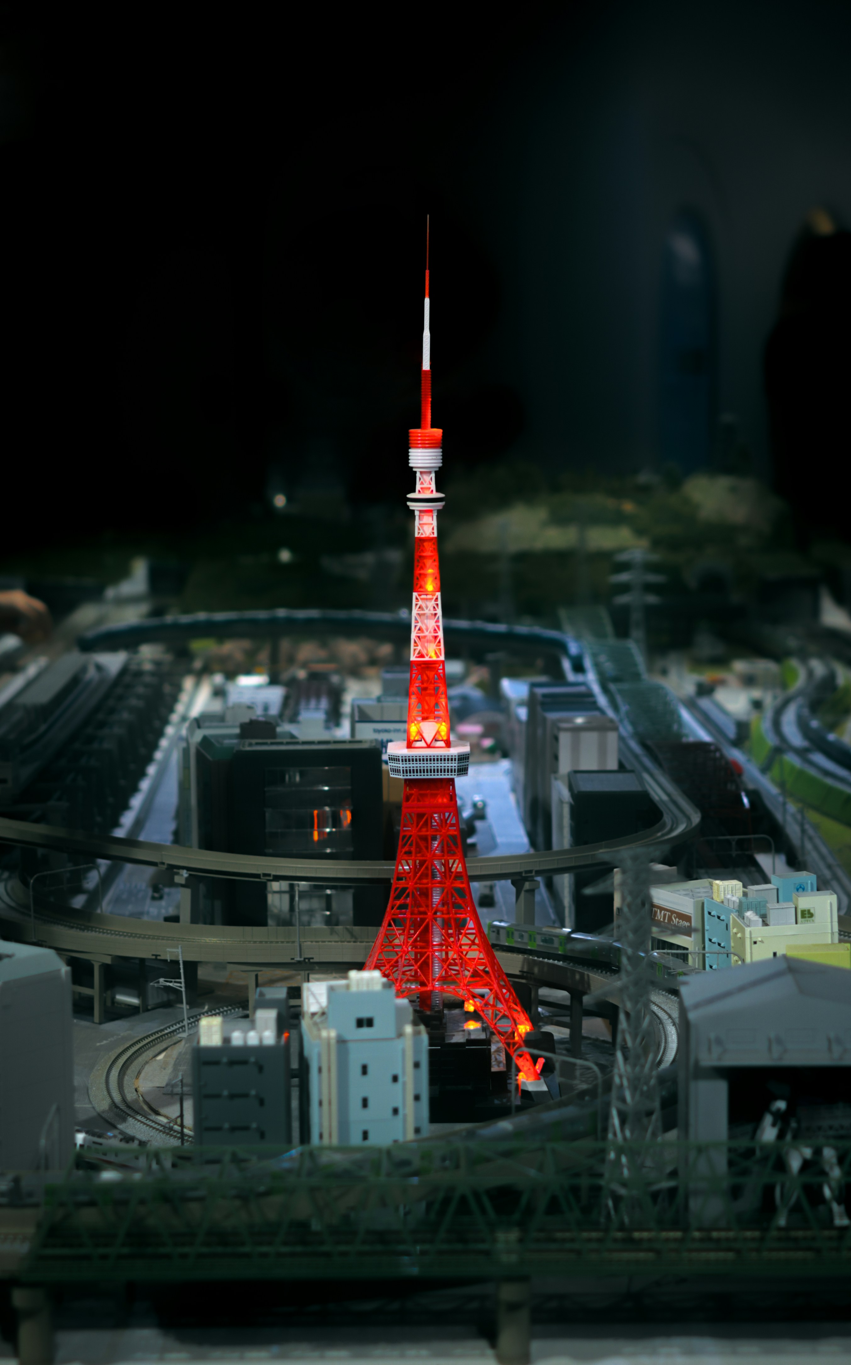 A detailed scale model of Tokyo Tower stands illuminated amid a miniature cityscape, with elevated tracks curving around the base.