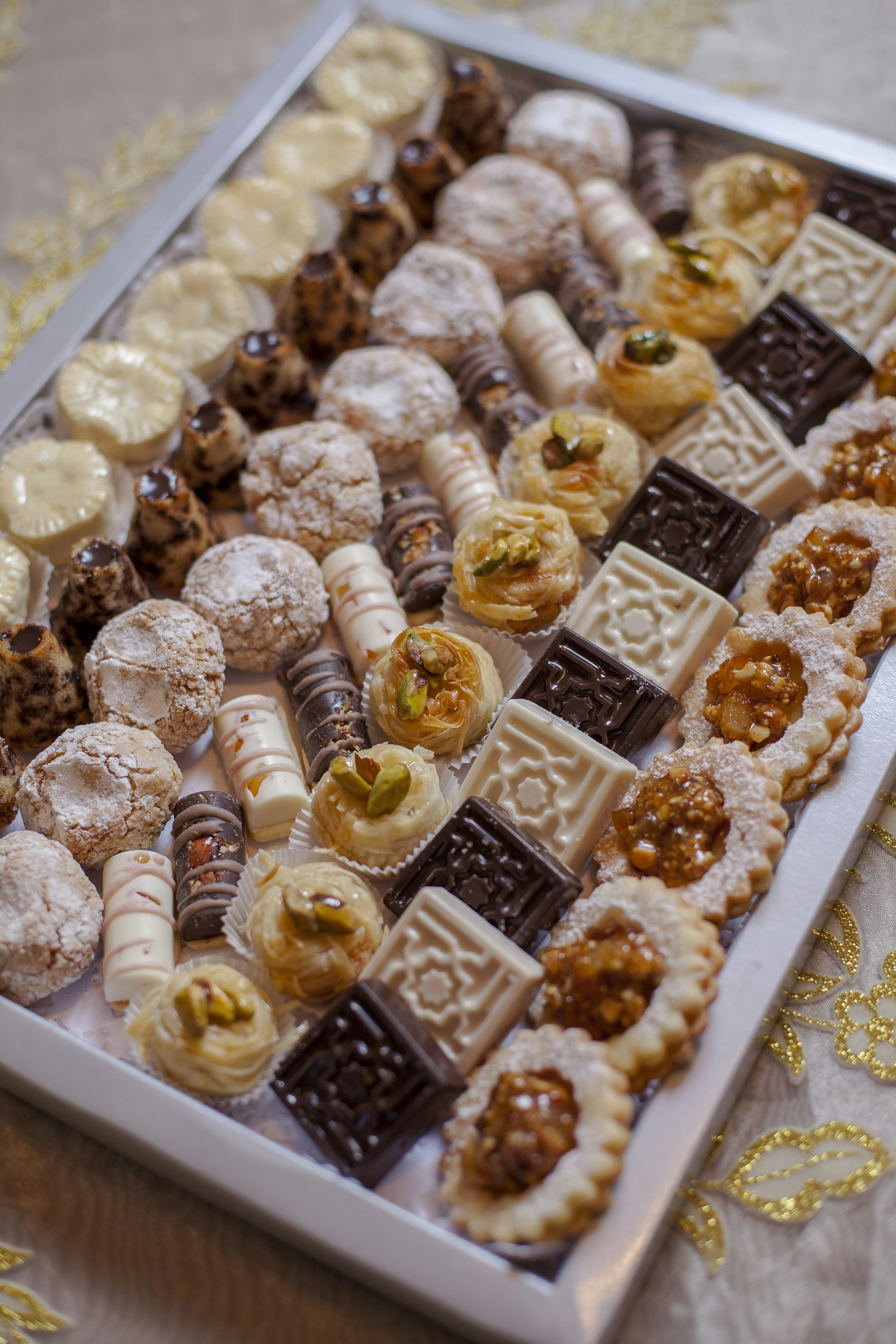 A glossy tray of assorted pastries and chocolates arranged in neat rows, featuring pistachio-topped tarts, chocolate squares, and round pastries dusted with sugar. A confectionery platter ready for serving.