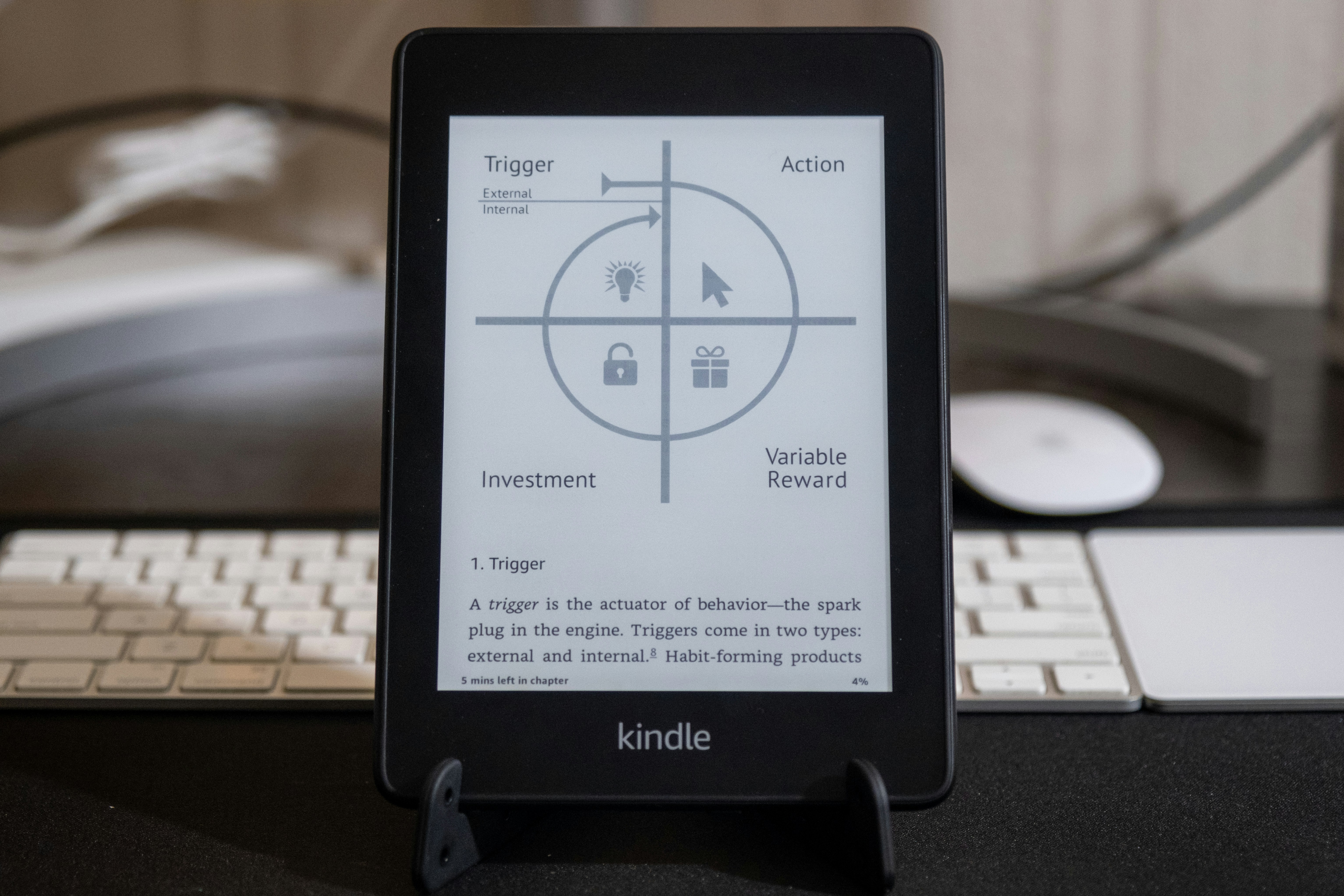 An Amazon Kindle device displayed on a small stand, centered on a desk with a keyboard and blurred background, signaling a focused reading/tech setup. The setup includes a logo-bearing e-reader framed by accessories in a muted, productive workspace.