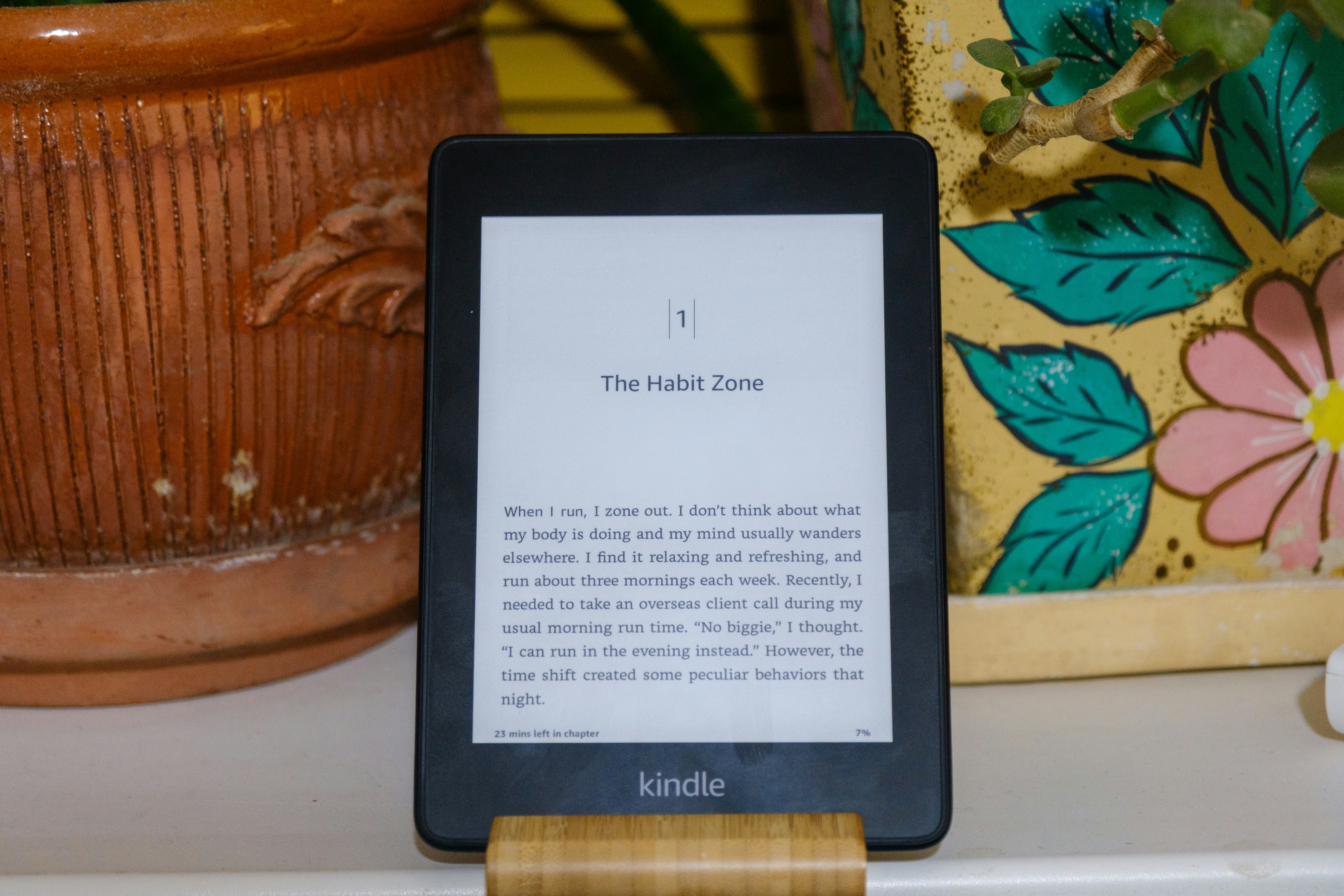A Kindle on a wooden stand rests between a terracotta pot and a colorful floral artwork, emphasizing the device as the calm focal point amidst textured backgrounds.