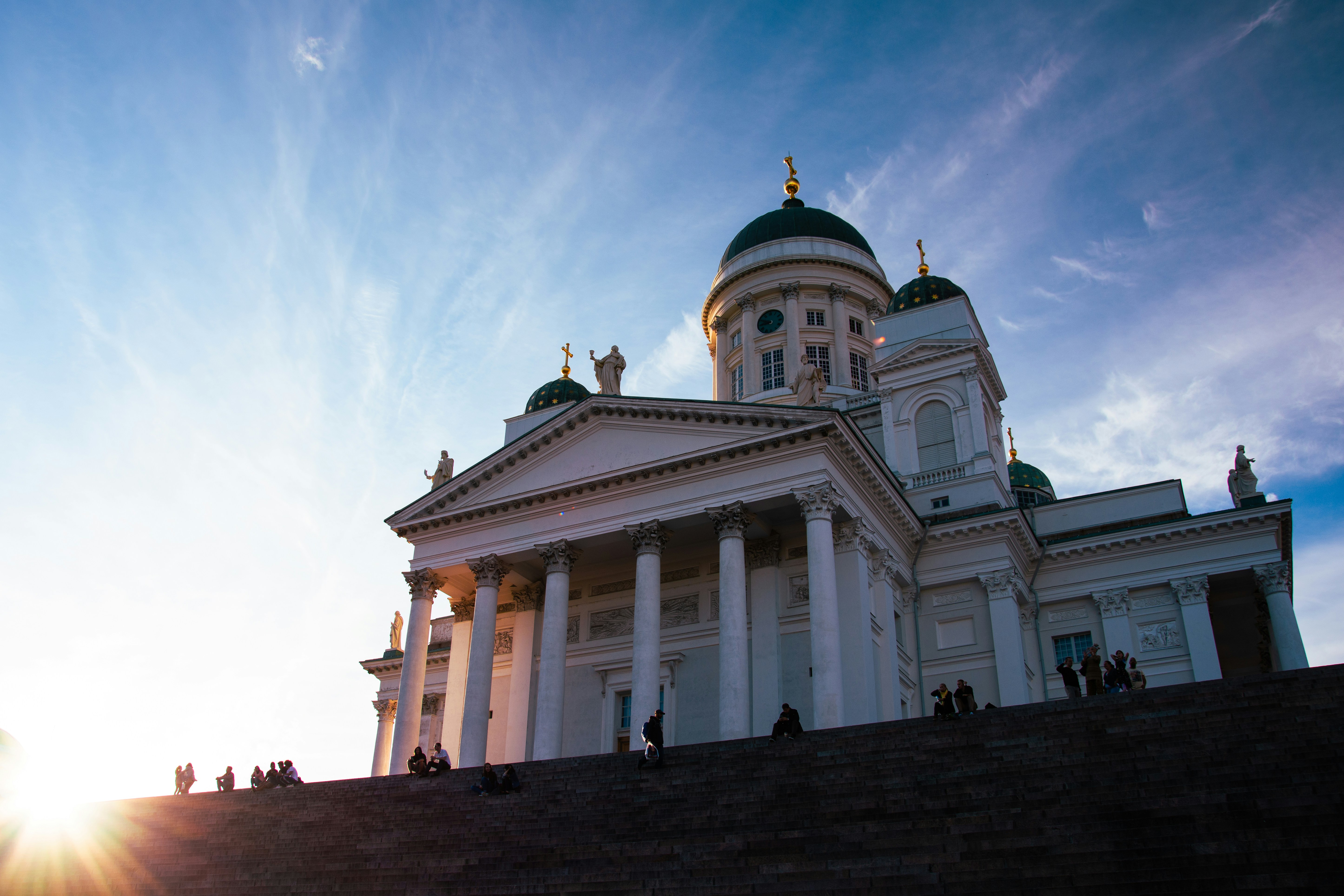 Sunlit neoclassical cathedral perched above broad stone steps with visitors along the façade; warm glow from the setting sun enhances the columns and domes.