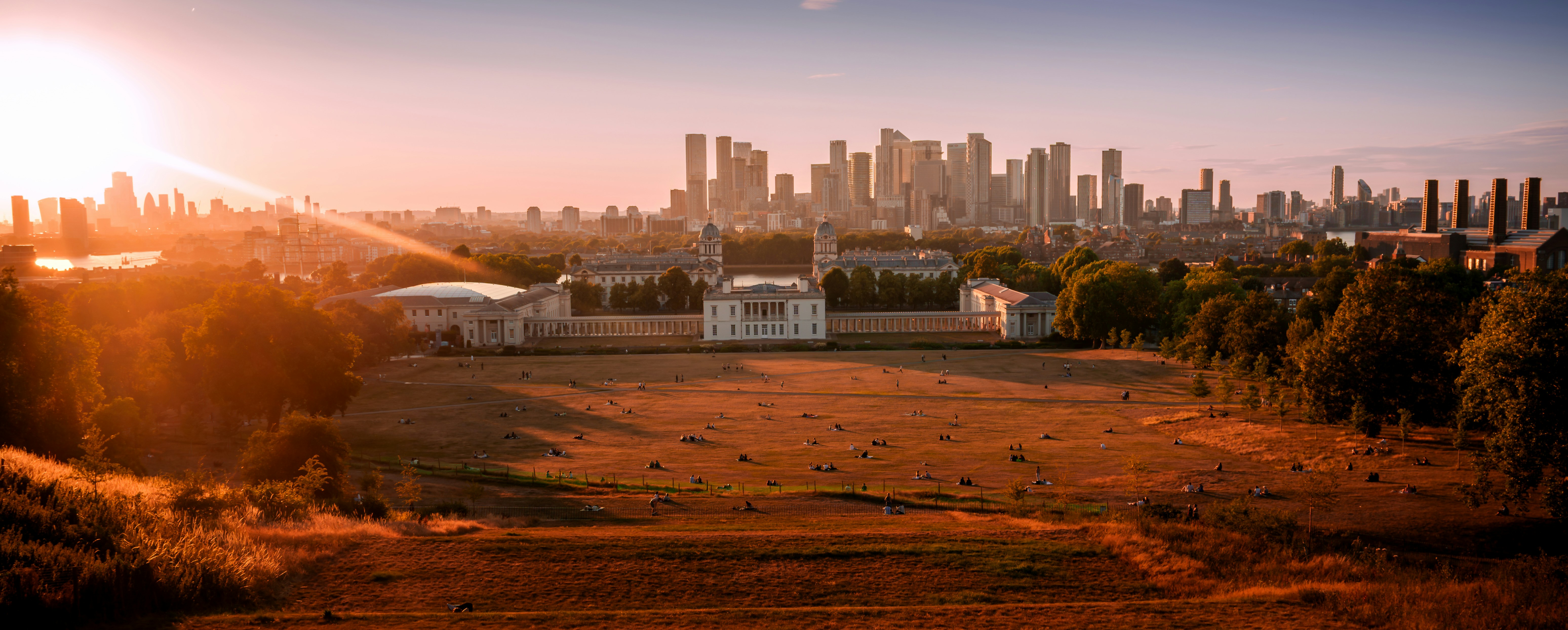Golden-hour landscape with a sprawling field and distant city skyline; the scene includes historic buildings near the foreground and a warm, glowing horizon.