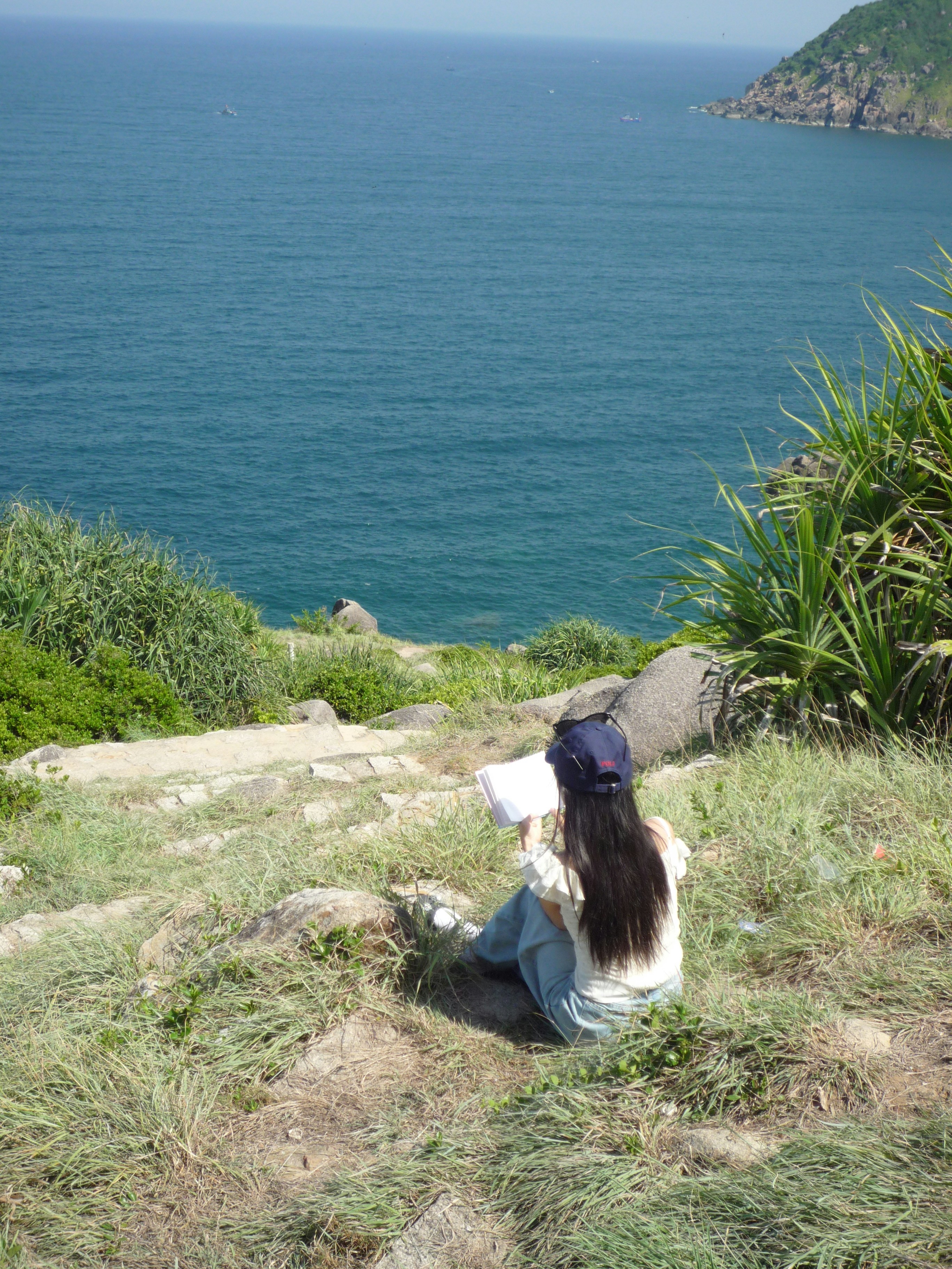 Woman sitting on a grassy cliff, gazing at the ocean.