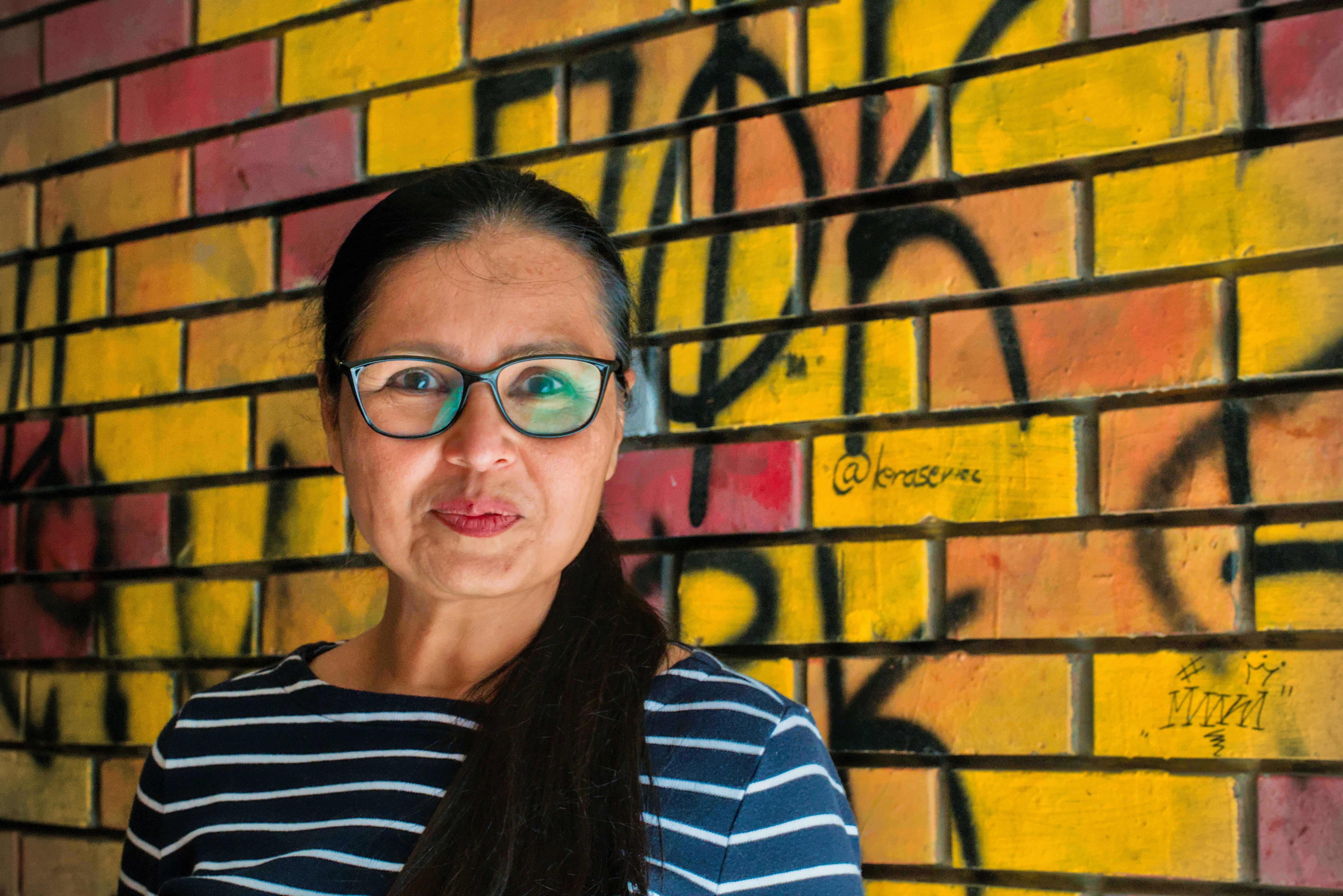 Portrait of a woman with blue glasses and a striped shirt against a vibrant graffiti wall, conveying a candid urban vibe.