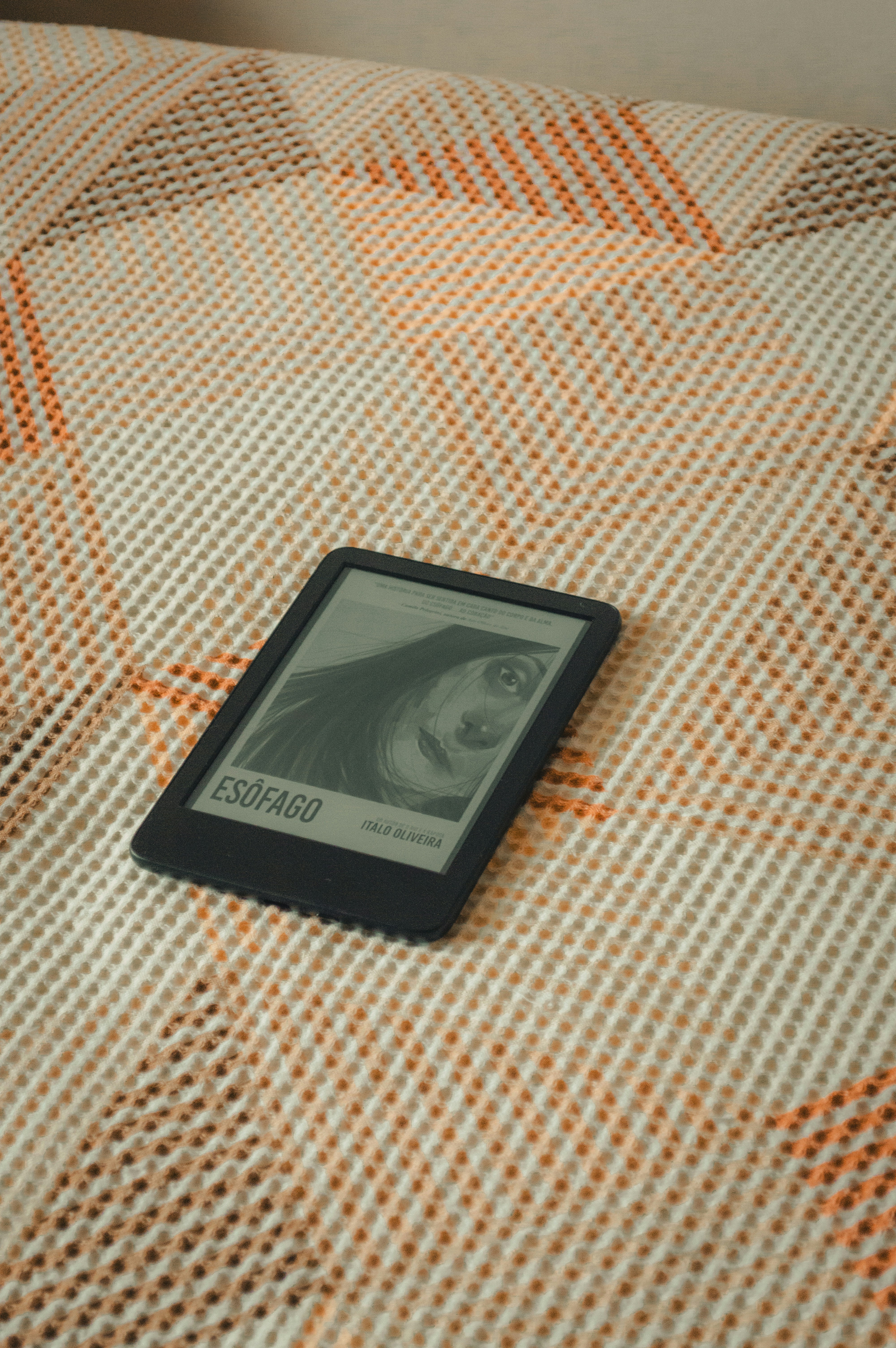 A small e-reader lies on a geometric, orange-and-cream quilt, displaying a grayscale screen with a visible image and text. The scene emphasizes texture and a retro vibe.
