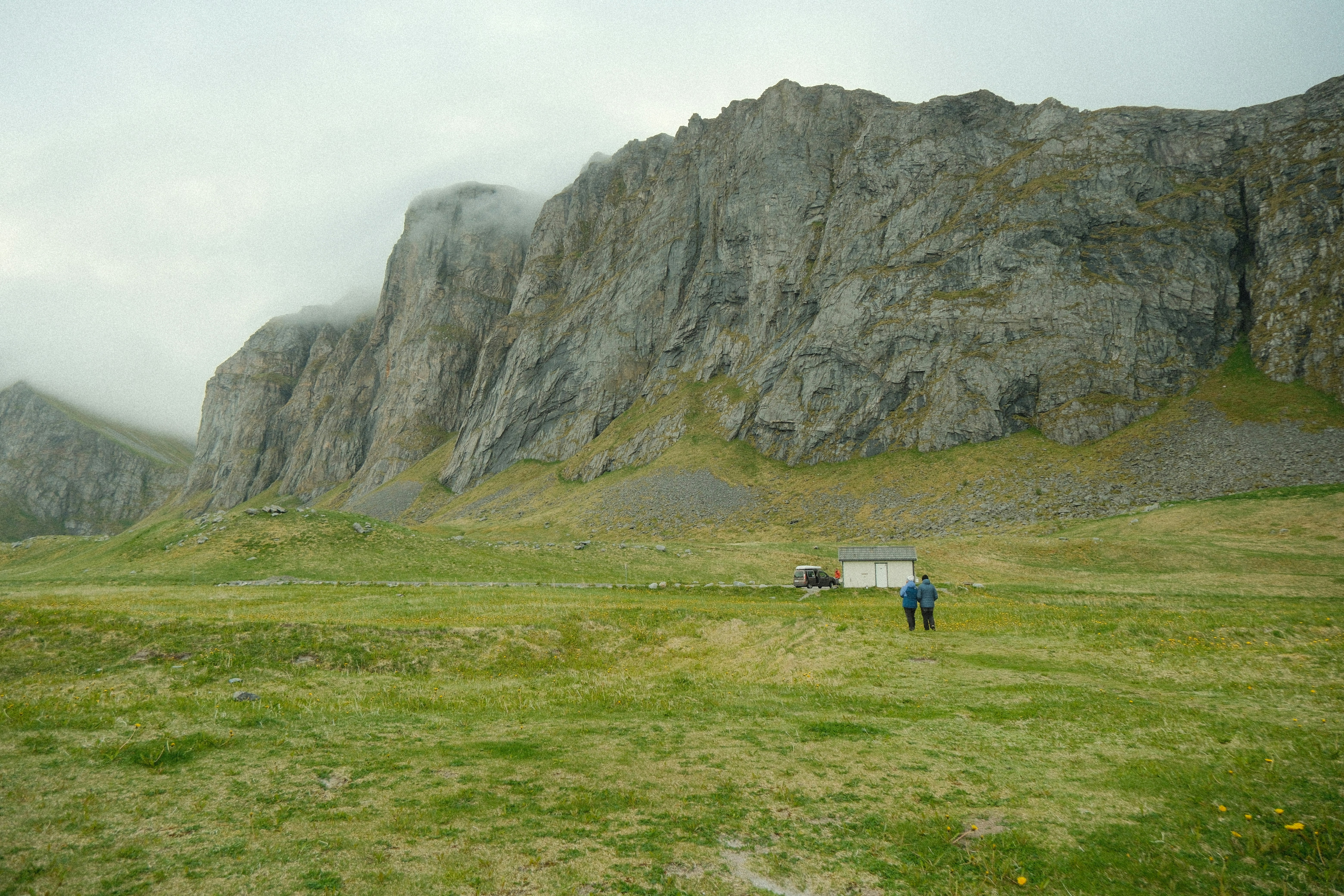 Two hikers at the base of a rugged cliff under a low grey sky