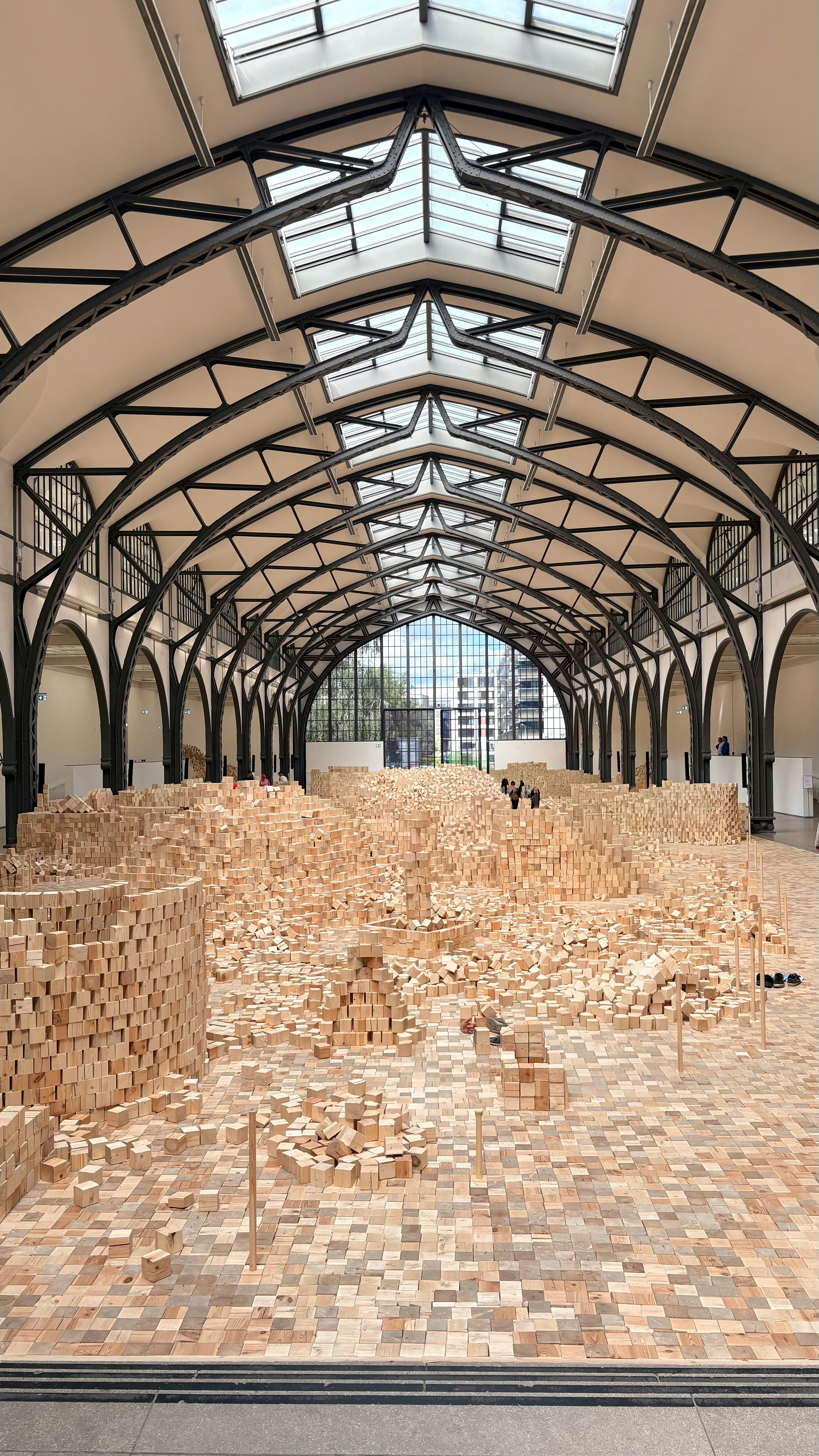 Massive timber block sculpture installation fills a grand train-hall-like space with arched steel beams and skylights; the wooden staggered forms extend toward a distant cityscape visible through the far end. This is a structured, large-scale sculpture/installation in a public hall.