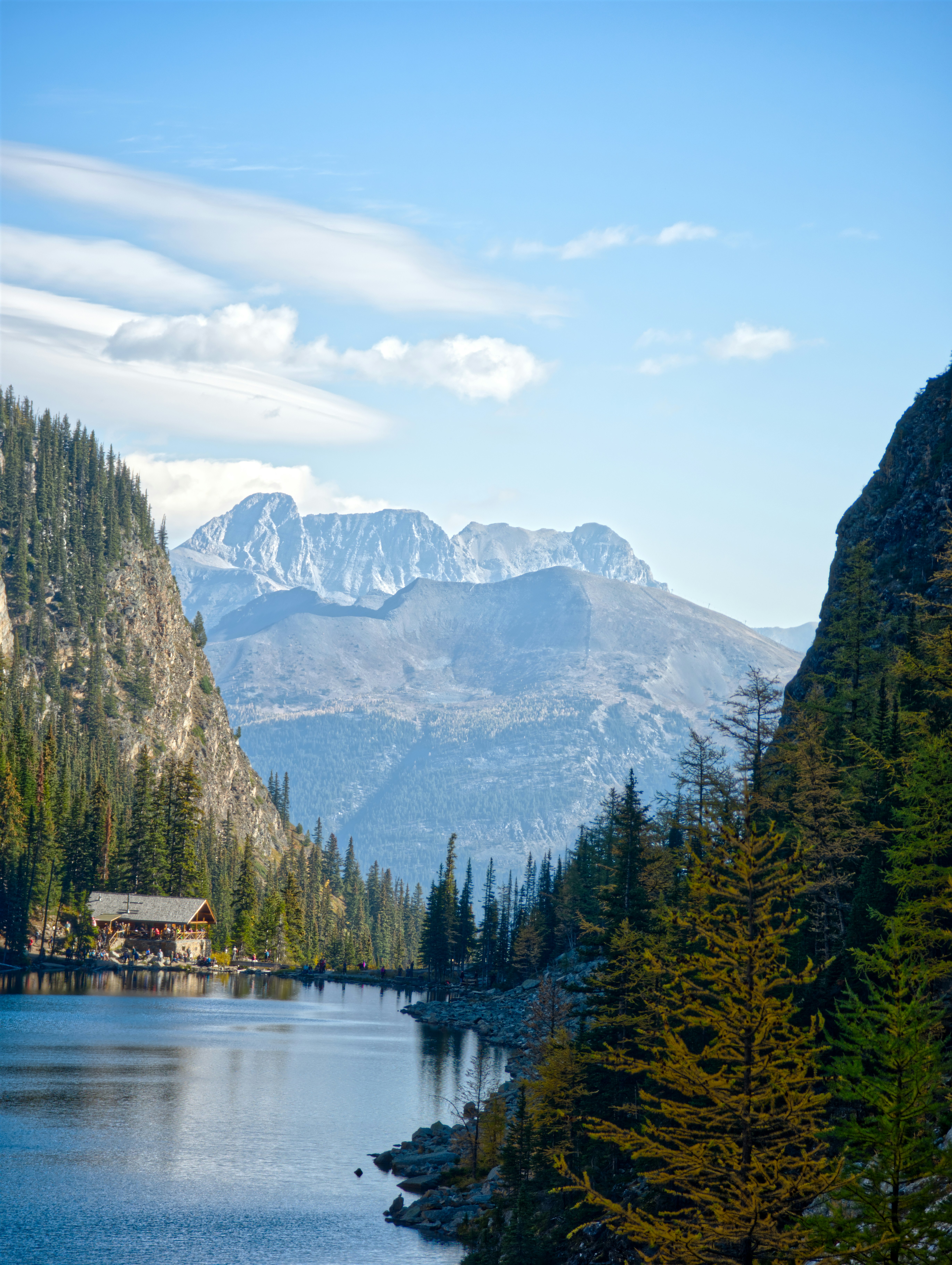 Cascading river valley with a calm reflective lake framed by forested slopes and towering mountains under a bright, clear sky. The scene emphasizes tranquil geometry and depth in a natural landscape.