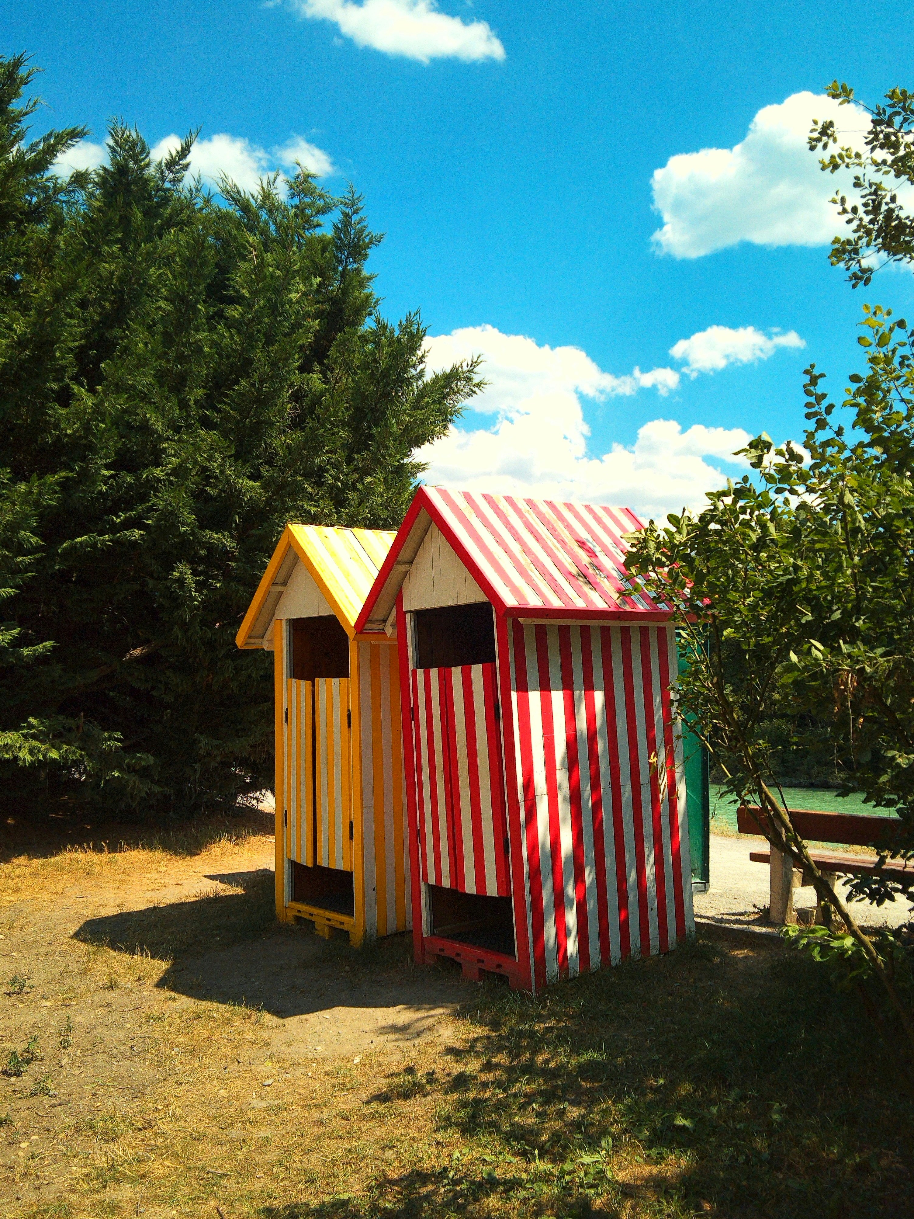 Bright striped beach huts stand side by side under a blue sky, casting long shadows on dry ground at the edge of a tree line.