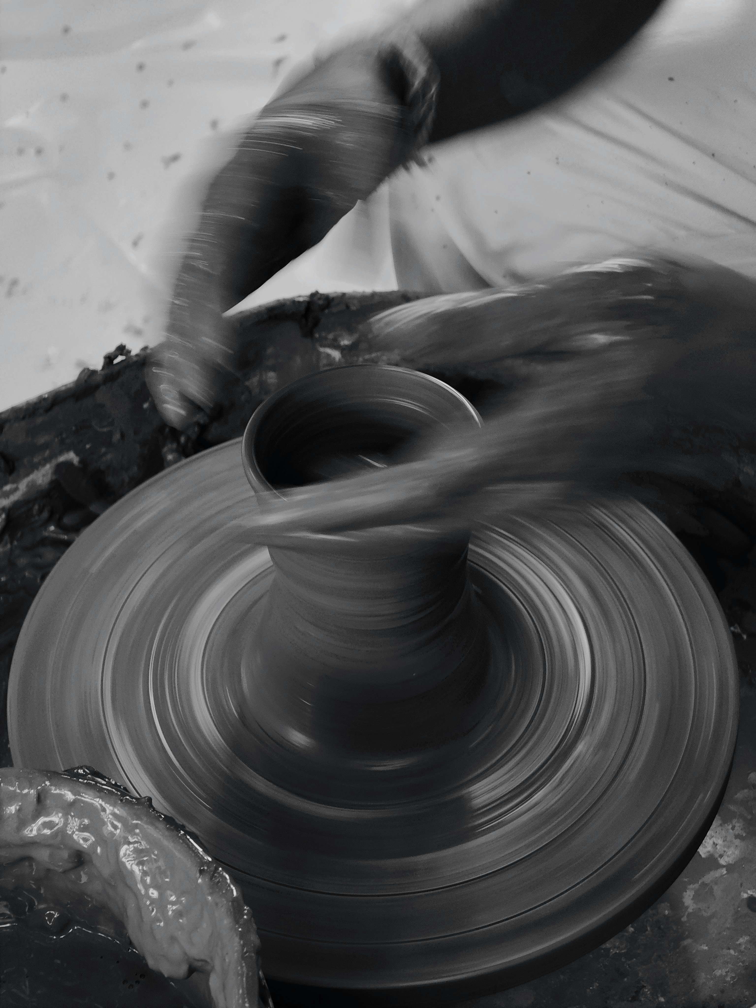 A grayscale close-up of a potter's wheel in action, with hands shaping a spinning clay vessel as the wheel blurs into motion.