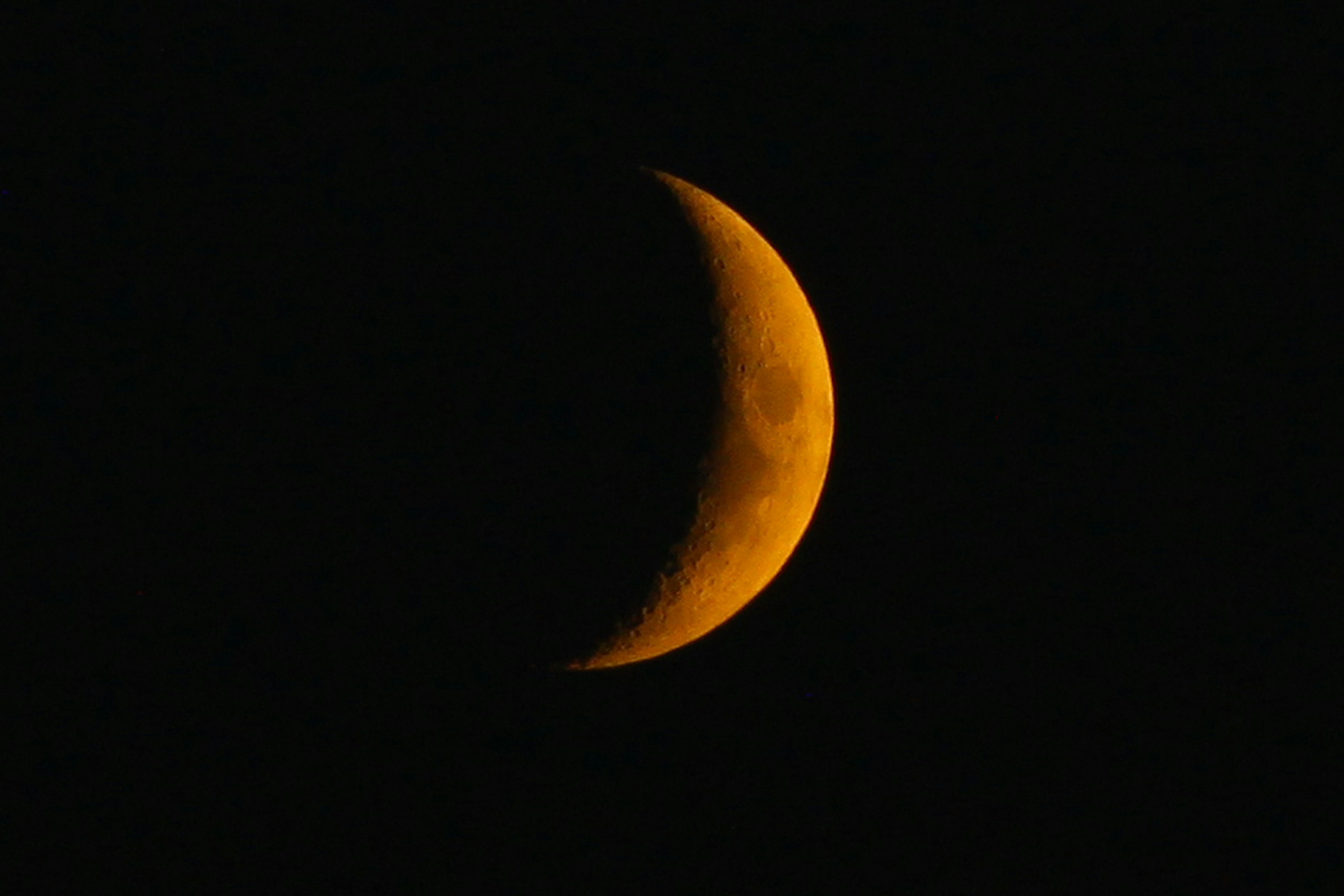 A thin, orange crescent dominates a dark sky, highlighting the sunlit edge of the Moon.
