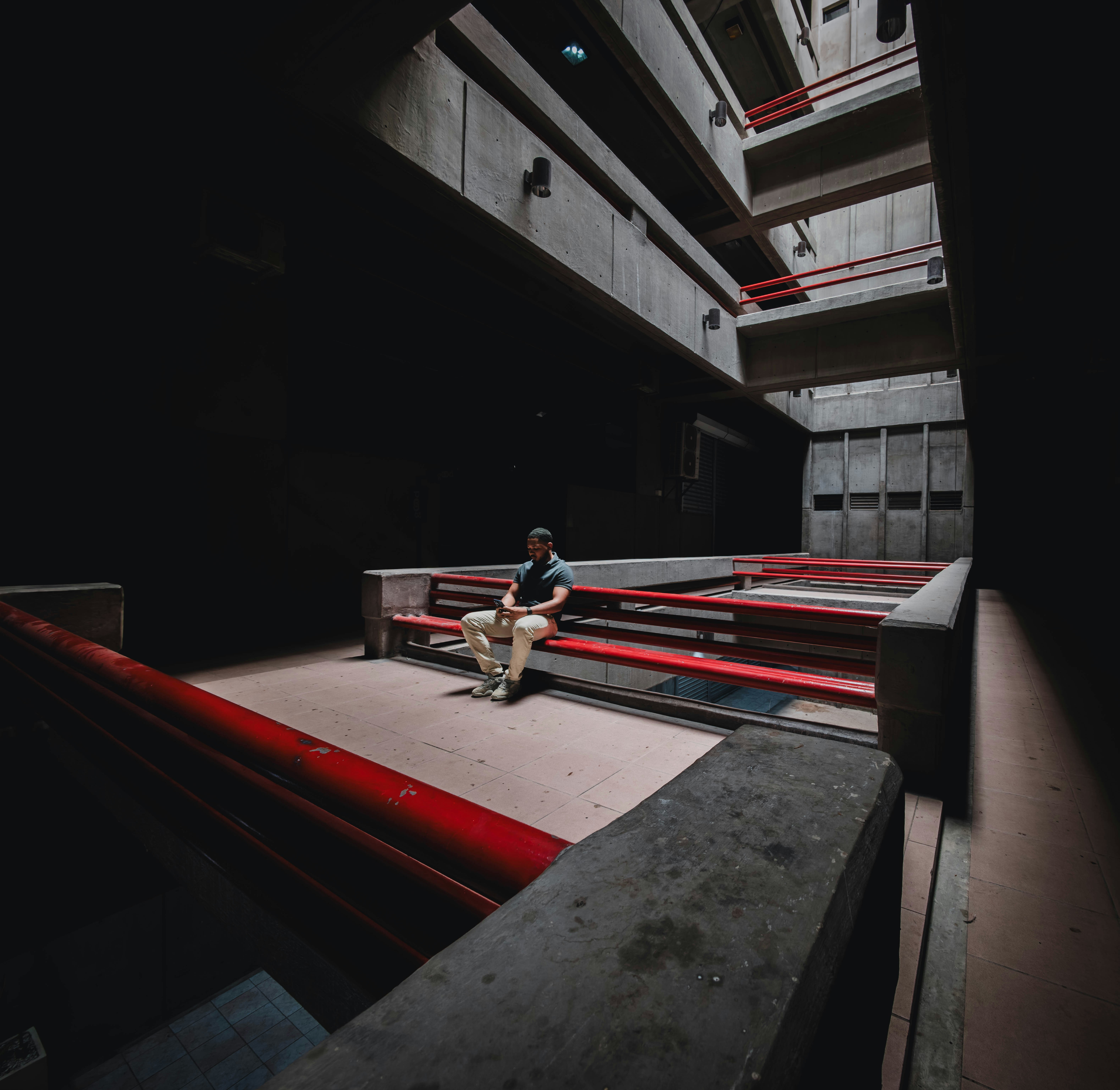 A lone person sits on bright red benches inside a stark, multi-story concrete atrium with dramatic lighting and perspective.