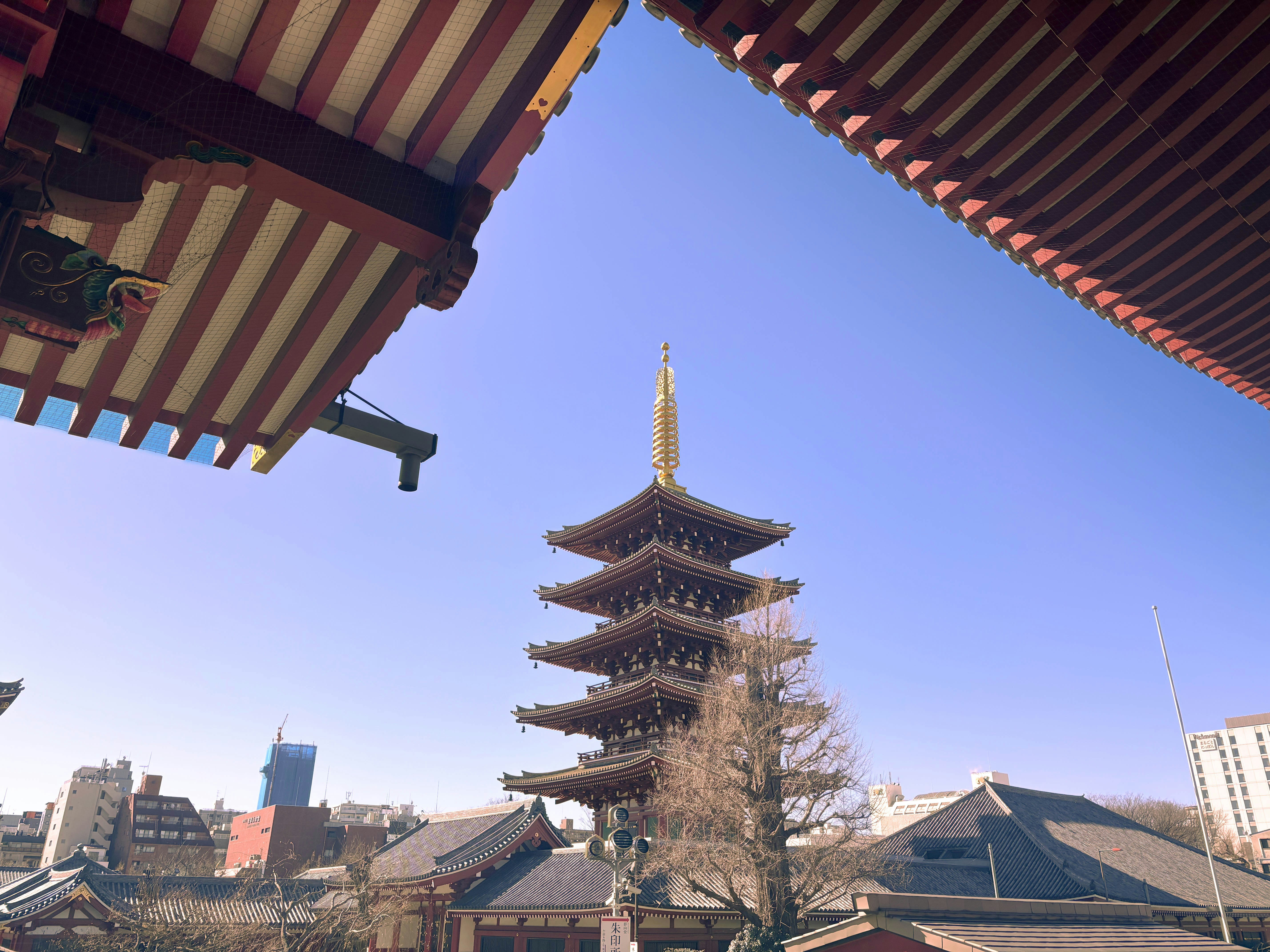 Temple complex with a central multi-story pagoda framed by temple eaves and urban backdrop, emphasizing traditional architecture. A clear blue sky and distant buildings provide context to the scene.