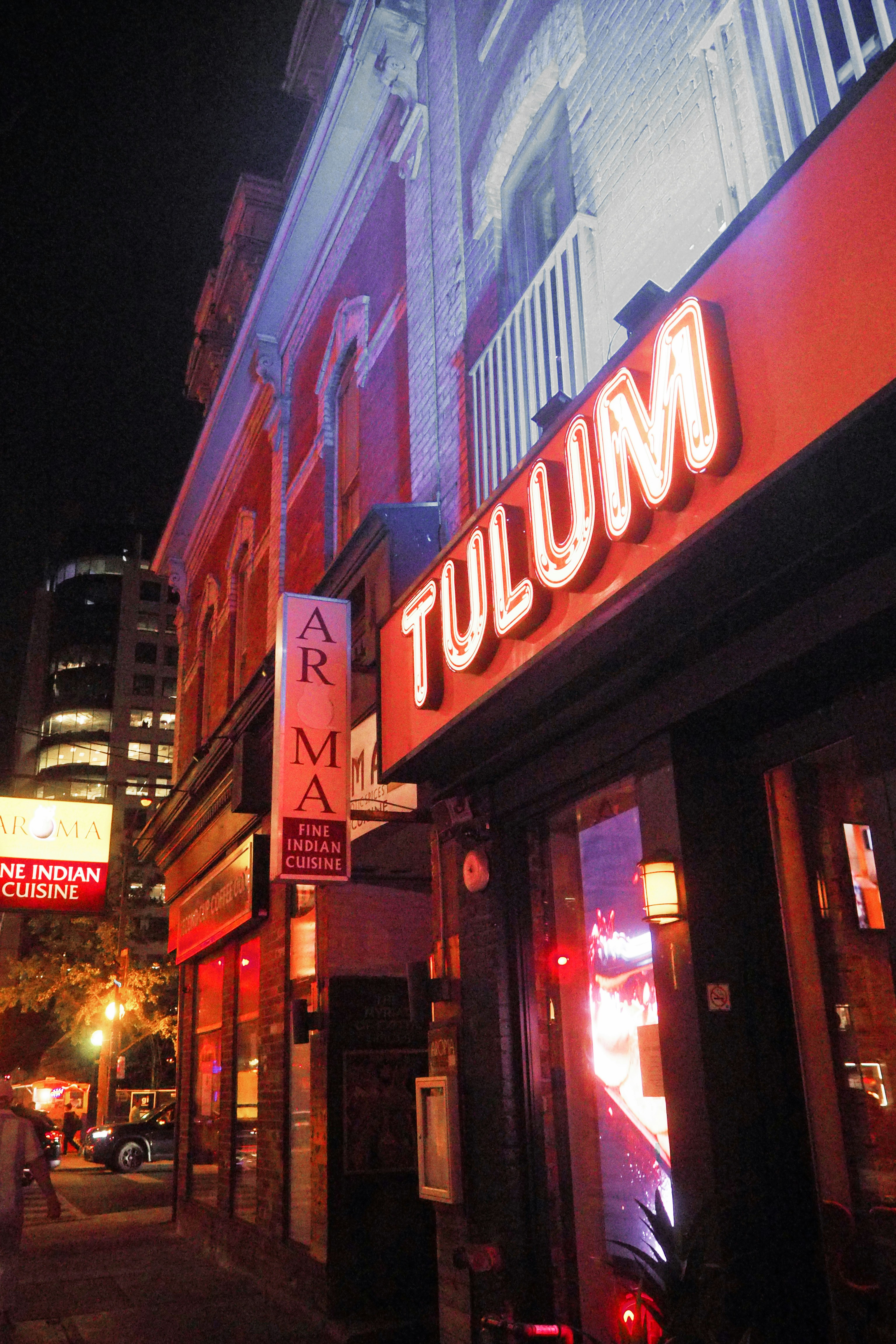 Bright neon sign and restaurant fronts line a city street at night, highlighting urban nightlife and Indian cuisine storefronts.