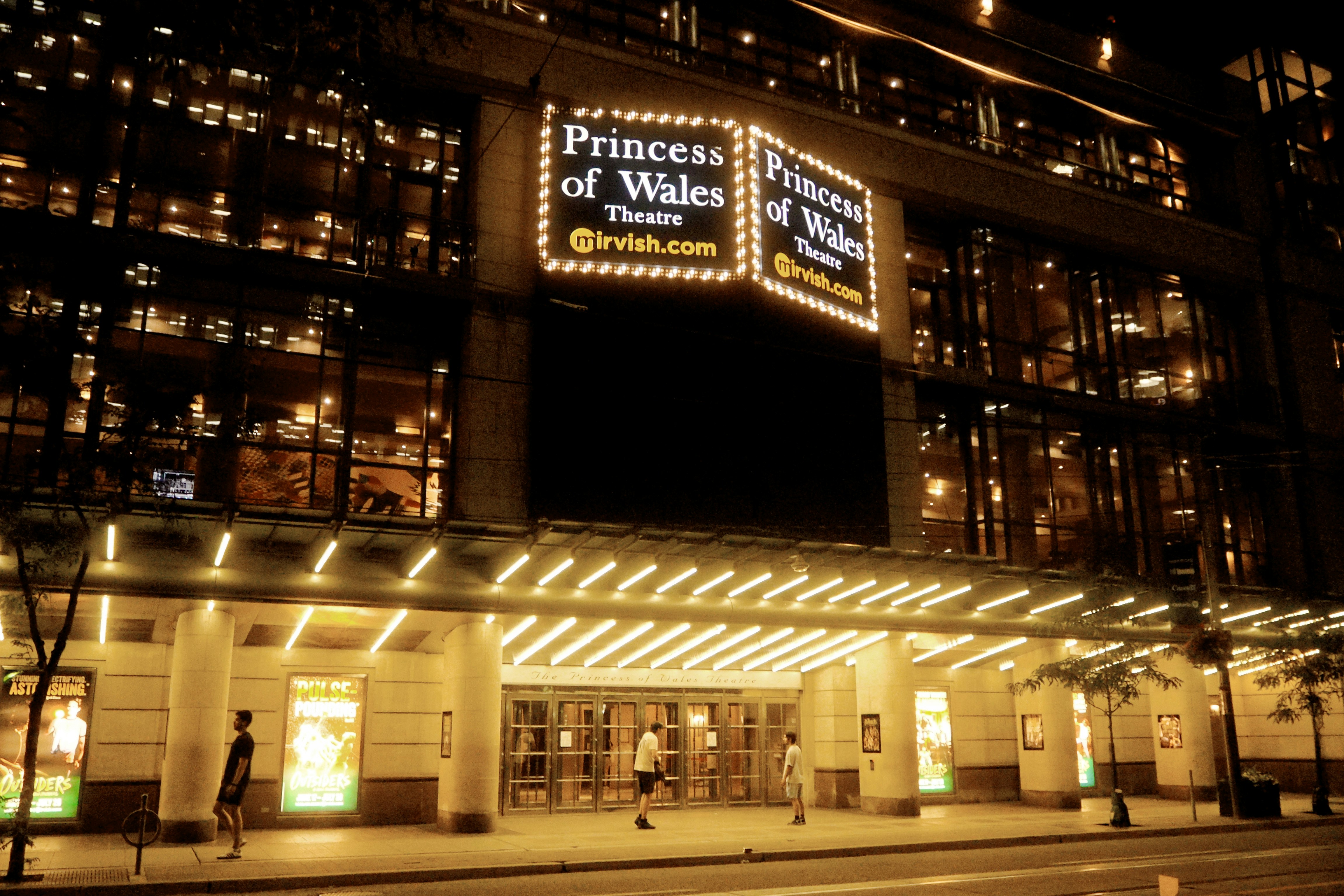 Warmly lit theater facade with marquee signs and glass-walled lobby, suggesting an evening show exterior.