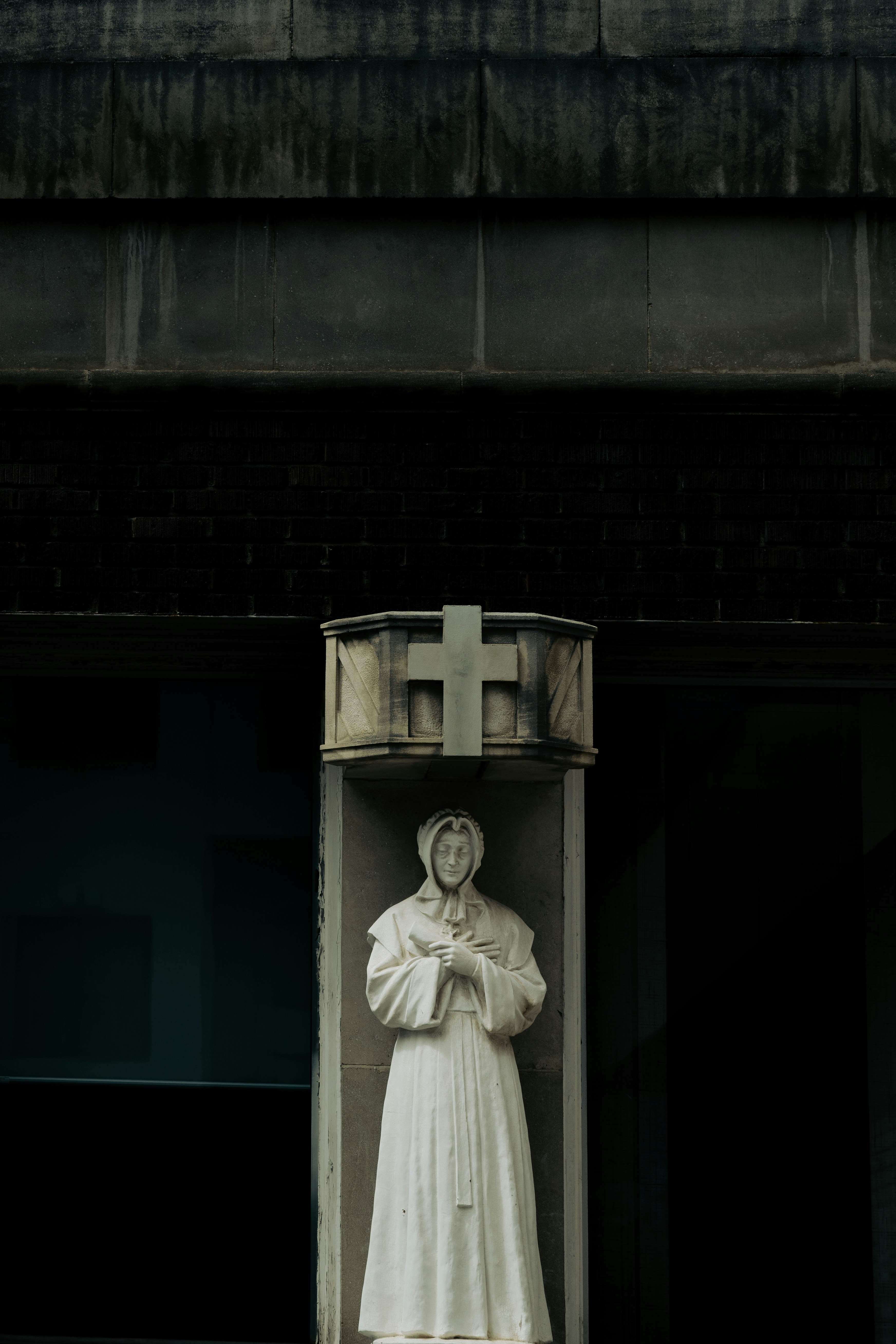 Monumental statue in a stone niche, hands crossed over chest, set against dark architectural backdrop; the composition emphasizes verticality and solemnity. Logo/monument photograph with subdued tones.