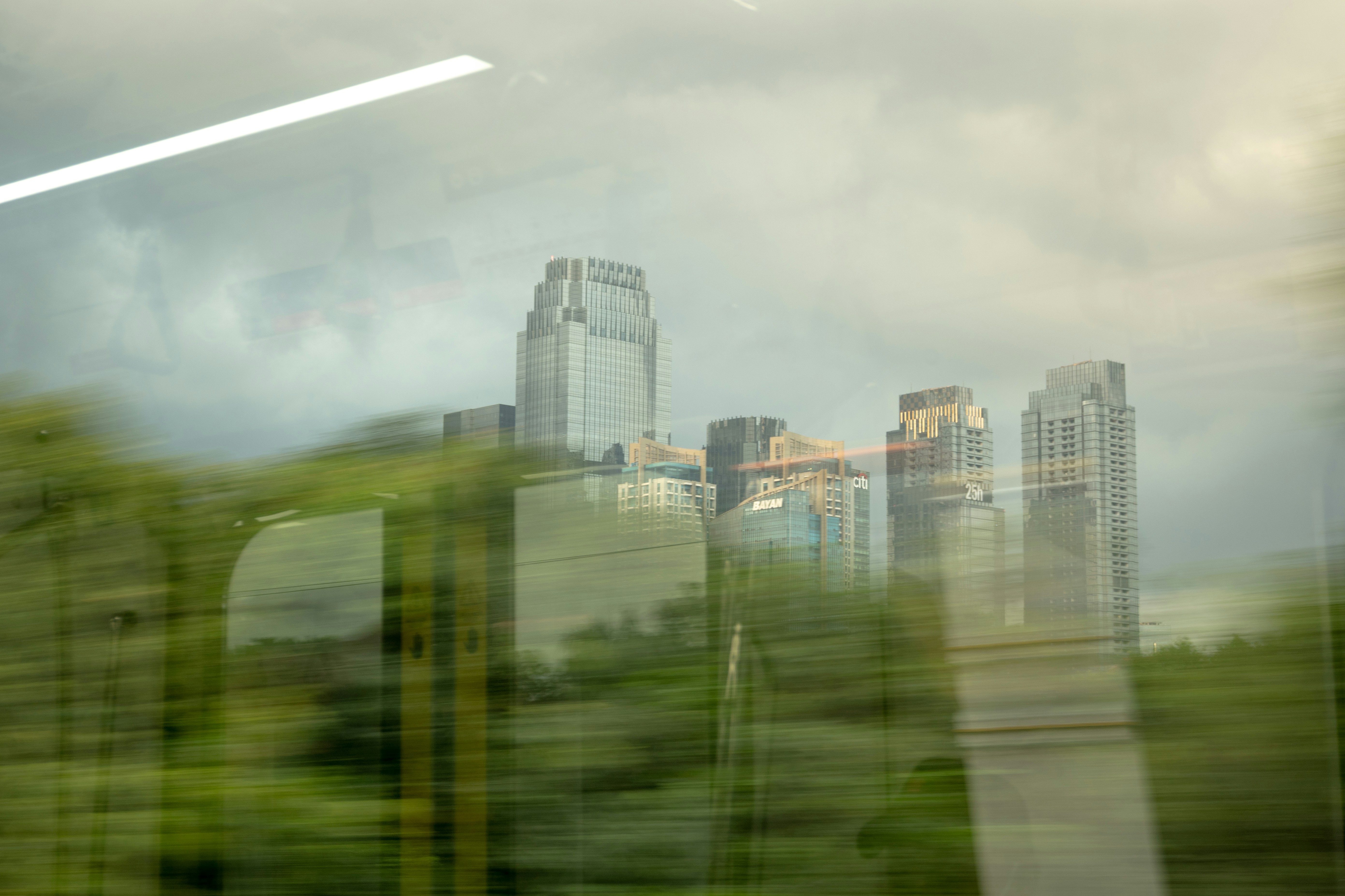 Blurry city skyline seen through a transit window with motion trails and reflections; a mix of high-rise buildings, greenery, and interior window elements. This is a photographic capture of movement and reflection.