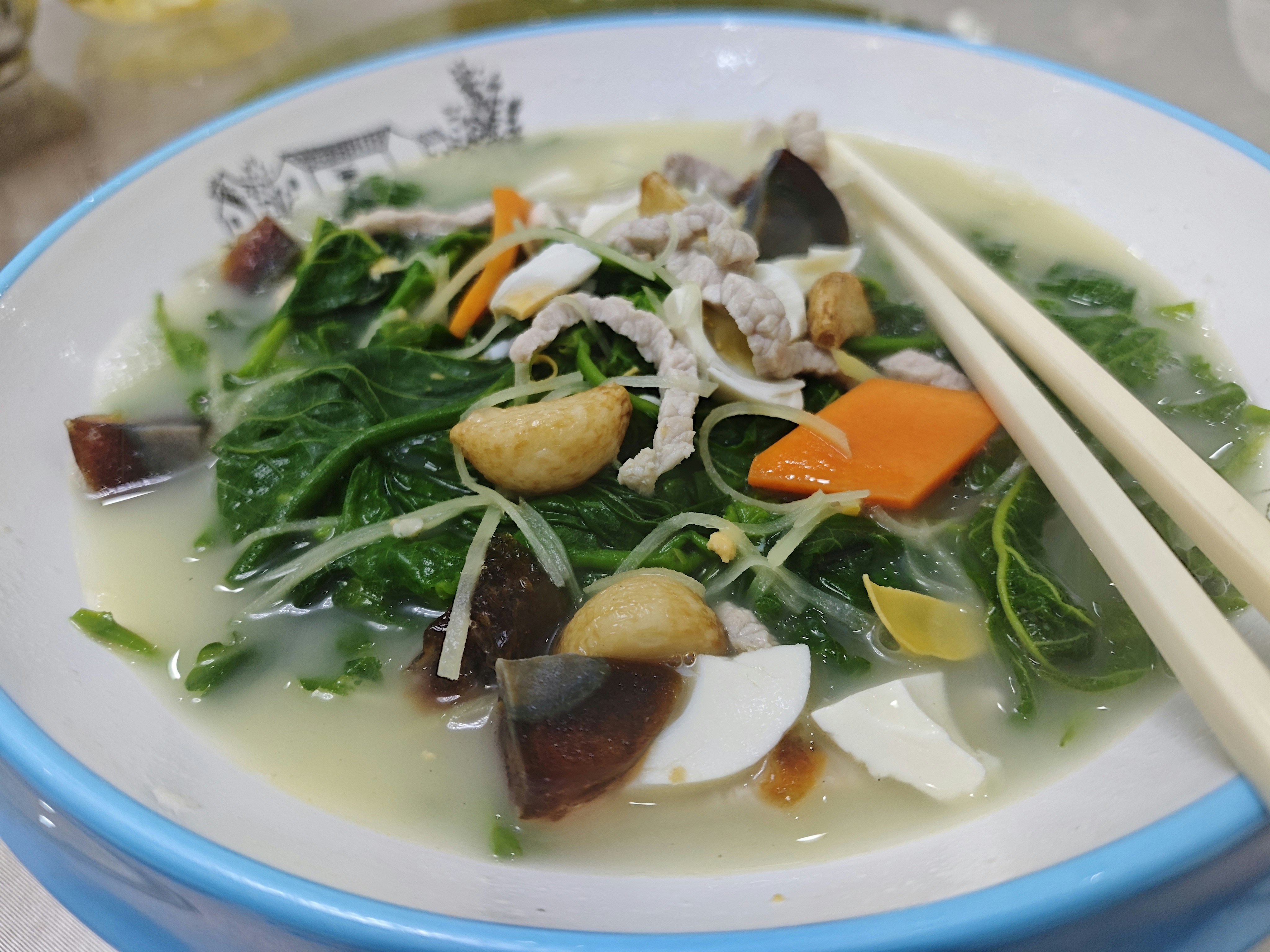Creamy green broth with leafy greens, tofu, and root vegetables in a blue-rimmed bowl, chopsticks resting on the edge; a nourishing Asian-inspired dish.
