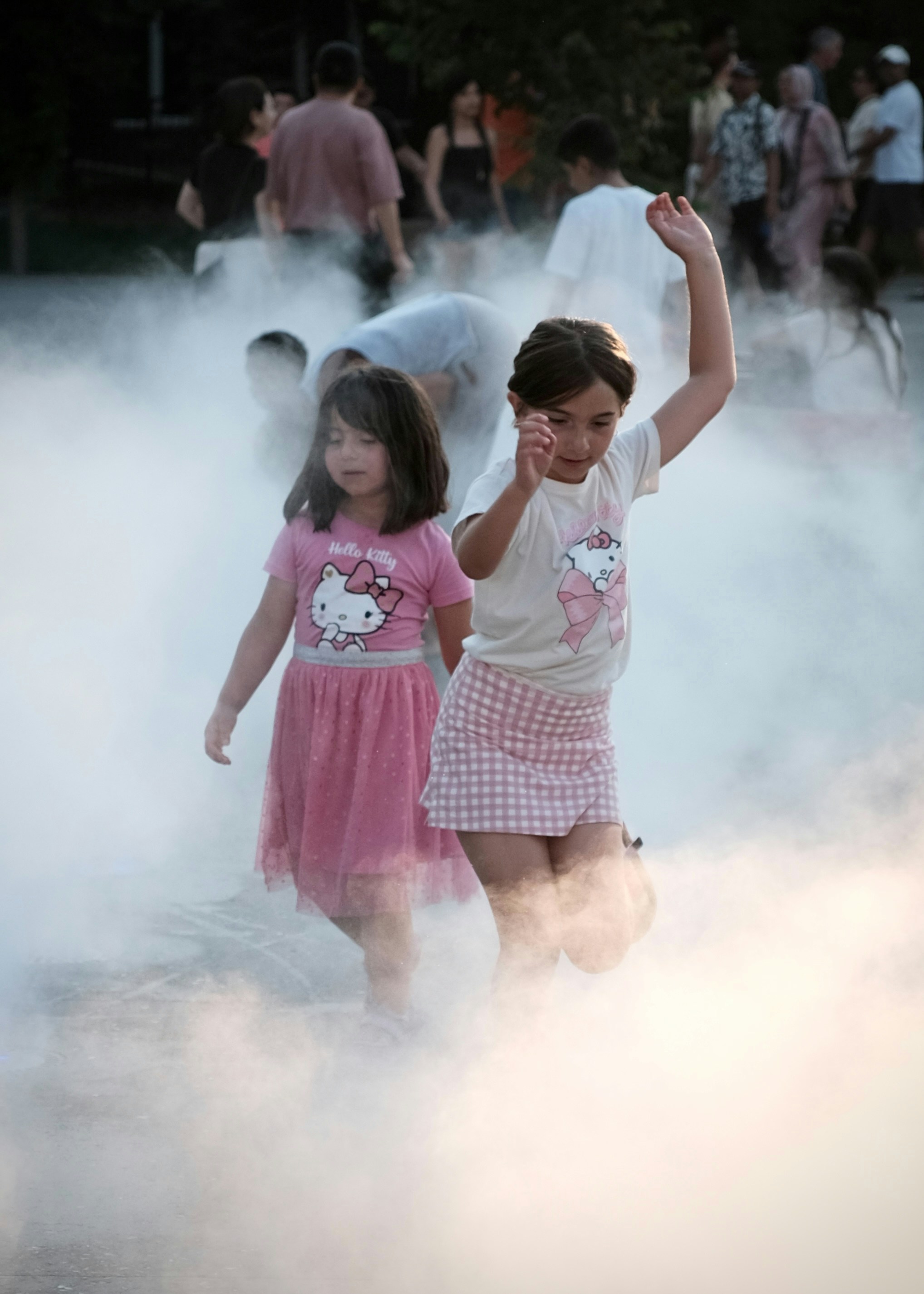 Girl in white shirt raises arm among fog with a pink-dressed companion at a smoky outdoor gathering