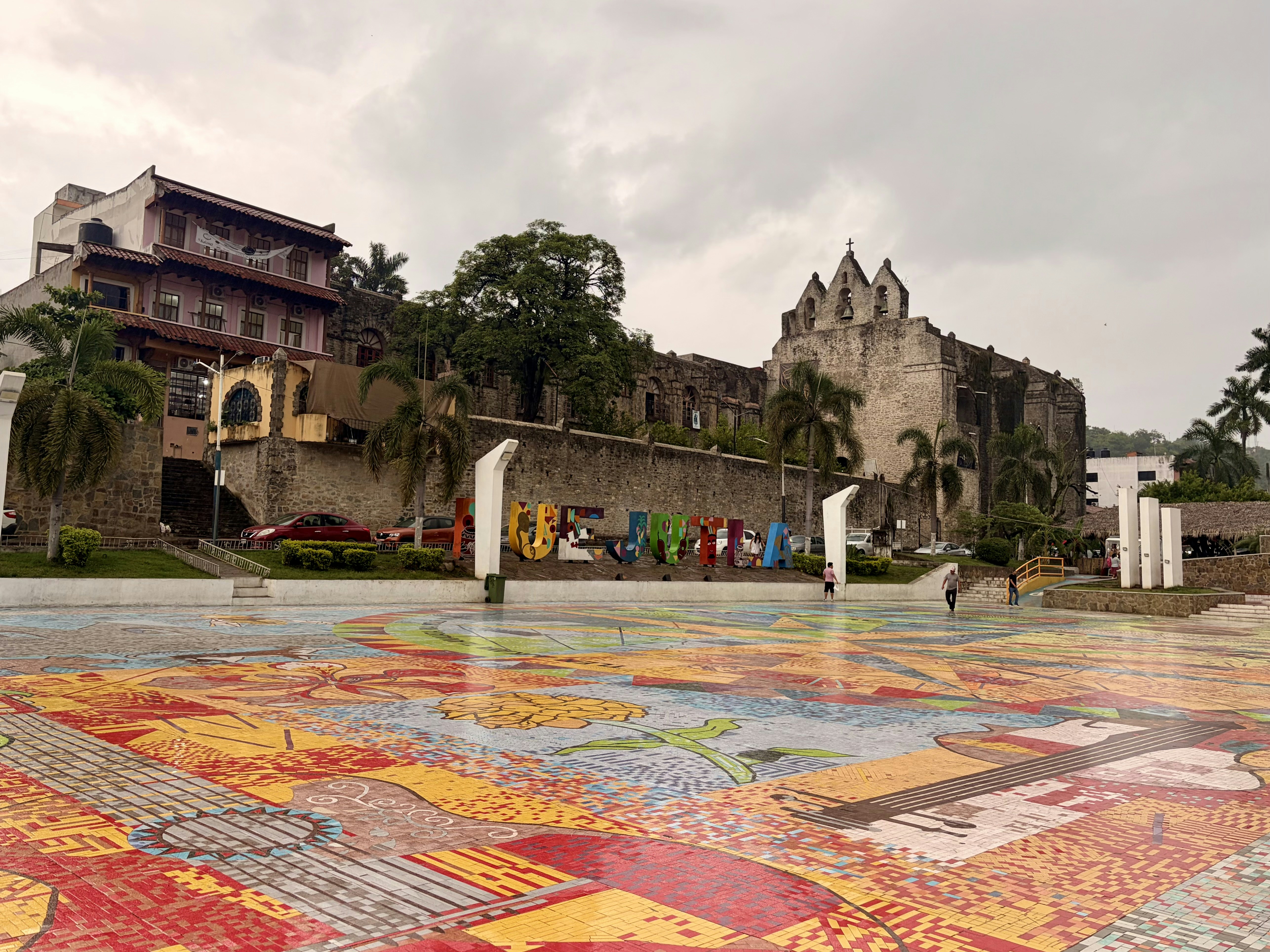 Vivid tiled plaza leading to a historic fort wall, with palm trees and a few visitors, foreground dominated by geometric mosaic patterns.