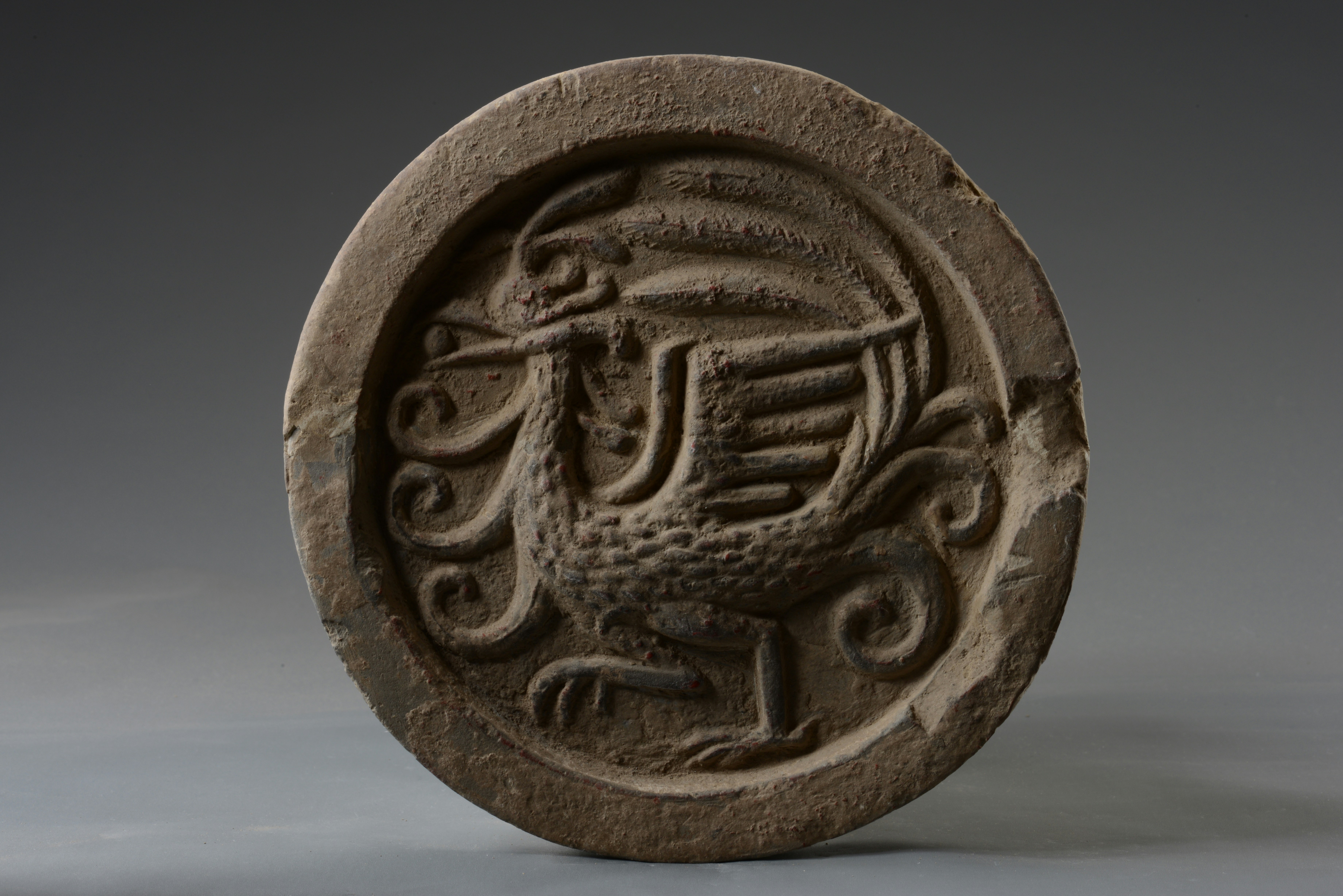 Round clay seal featuring a stylized bird emblem in relief, set against a smooth gradient backdrop. A pottery artifact showcasing intricate scrollwork and a weathered surface, presented as a study of texture and form.