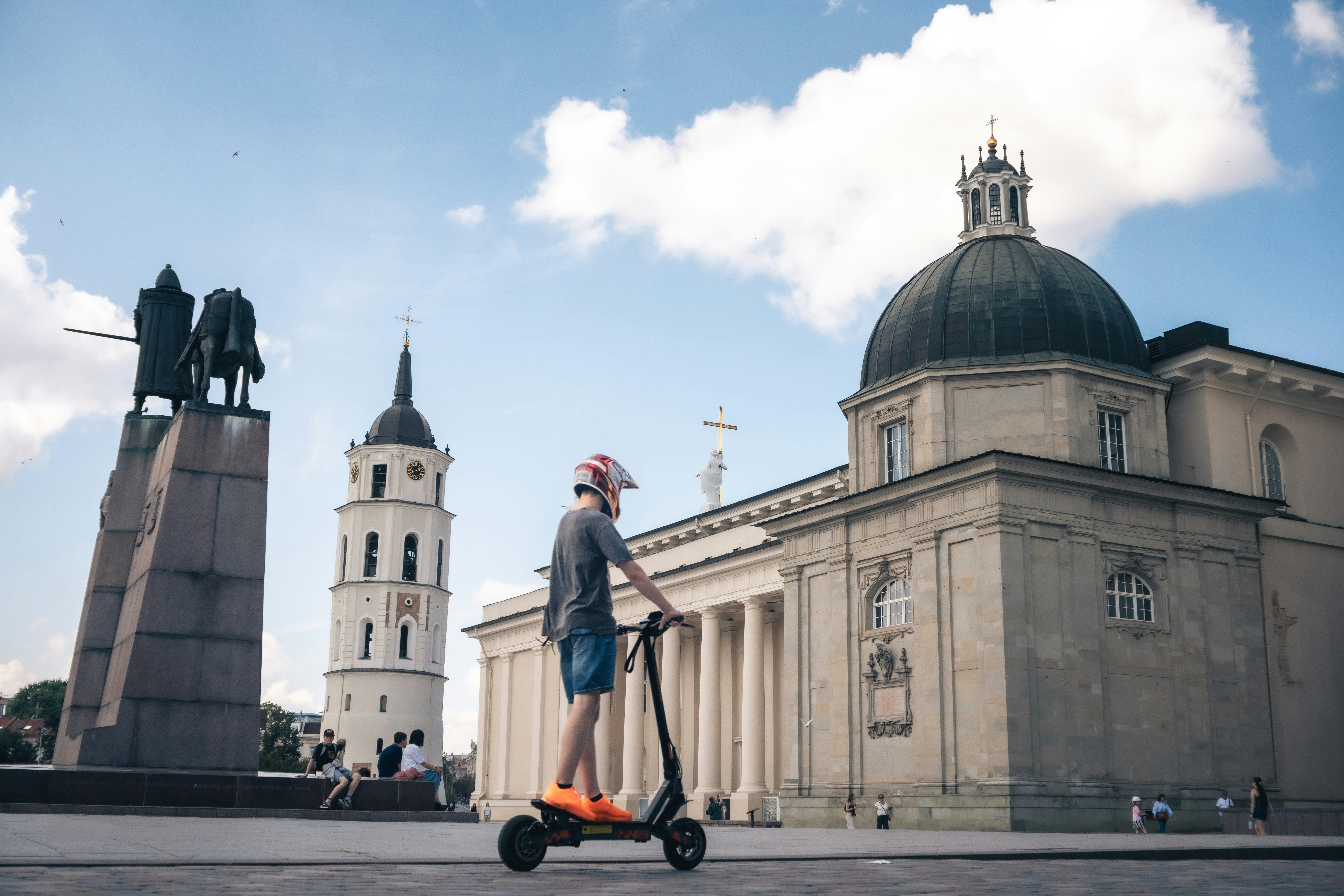 A rider on an electric scooter glides across a wide city square, with classical domed buildings and a statue in the background, under a bright daytime sky.