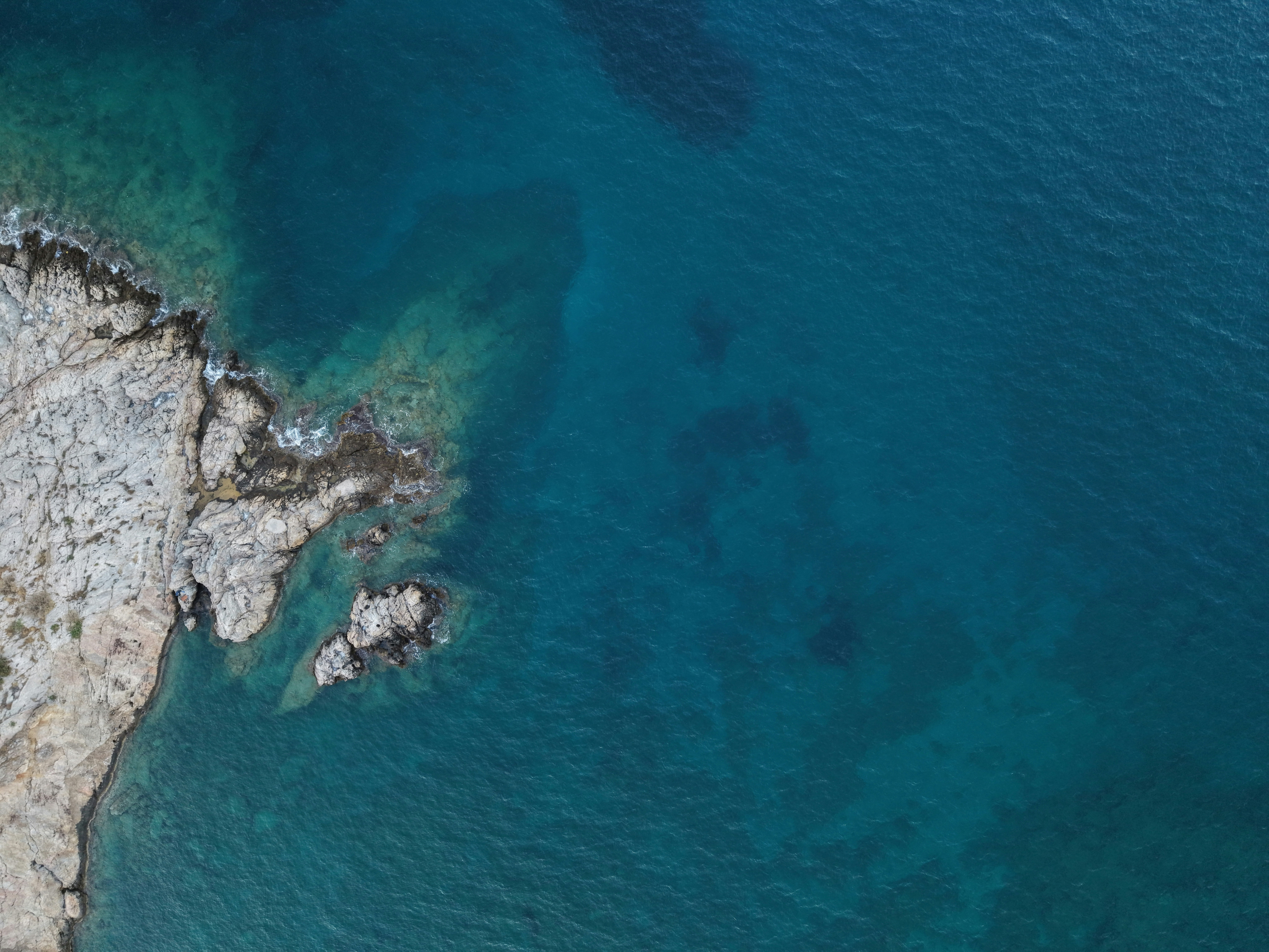 Aerial view of a rocky coastline where light turquoise waves meet rugged shoreline, revealing clear water gradients and submerged rocks.