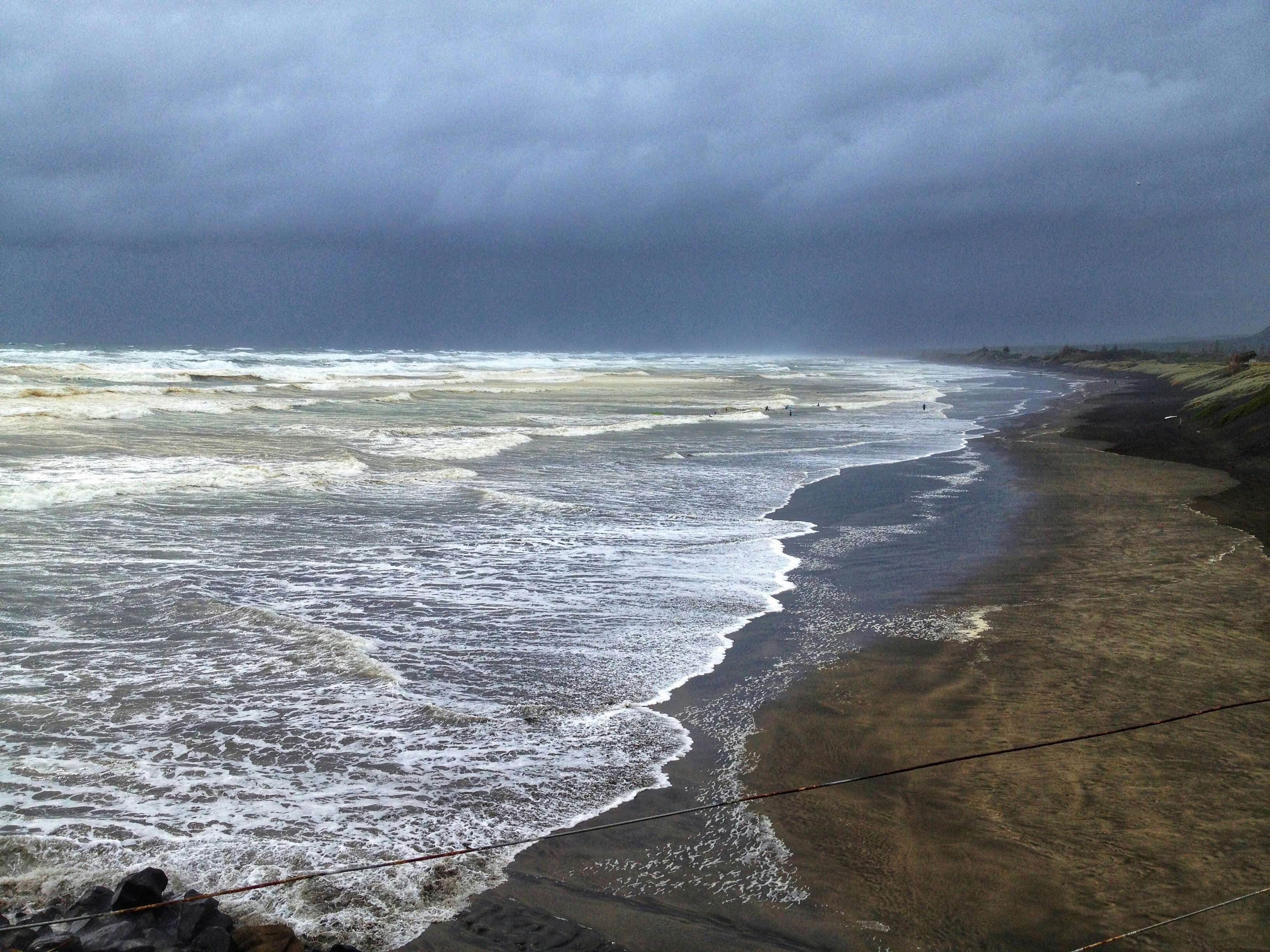 Rough sea along a windswept beach with breaking waves and a distant coastline under moody clouds.