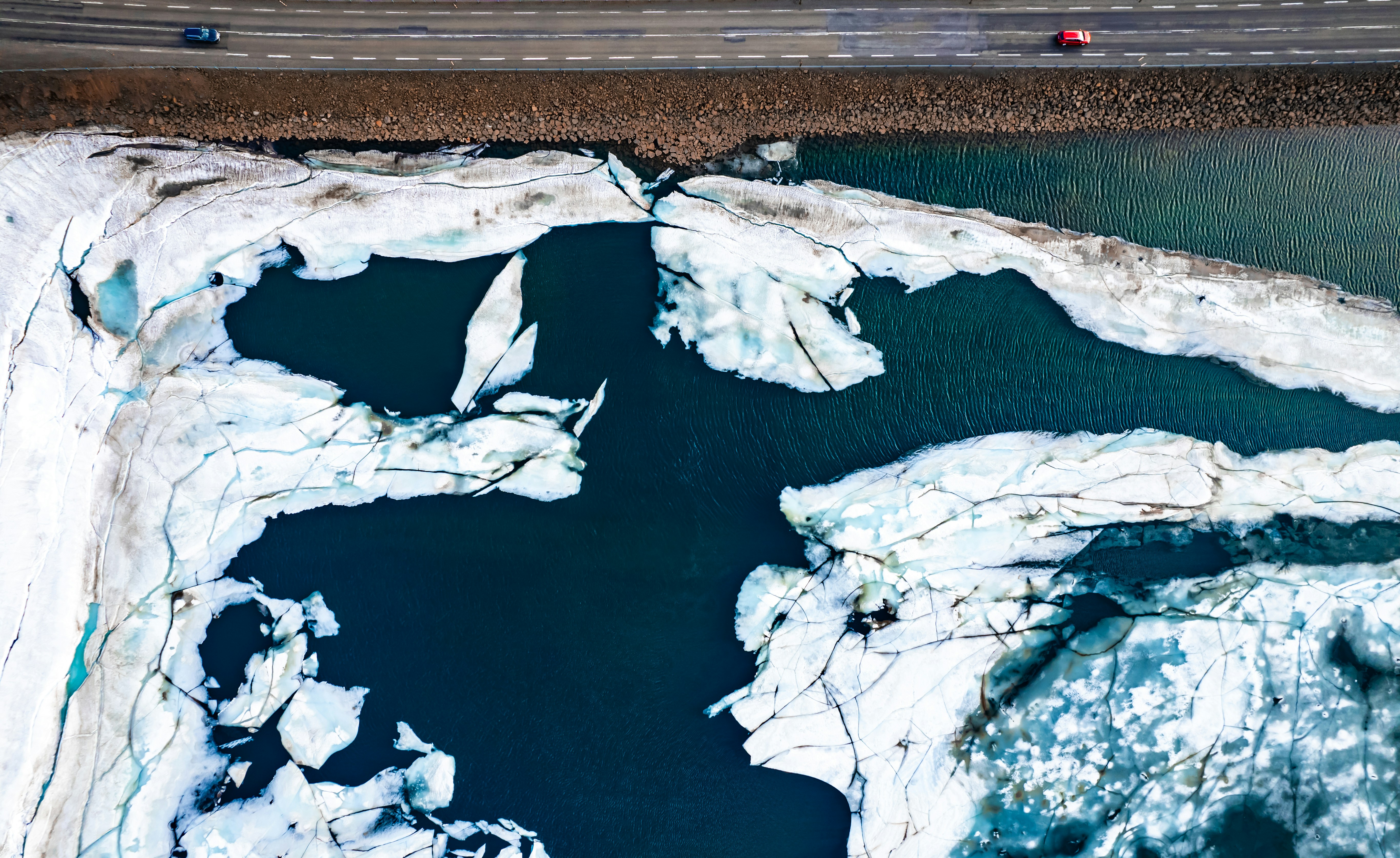Aerial view of a fractured ice field meeting dark blue water along a narrow channel, with a road running along the edge above. The scene emphasizes contrast between white ice and deep water, captured from above as a drone shot.