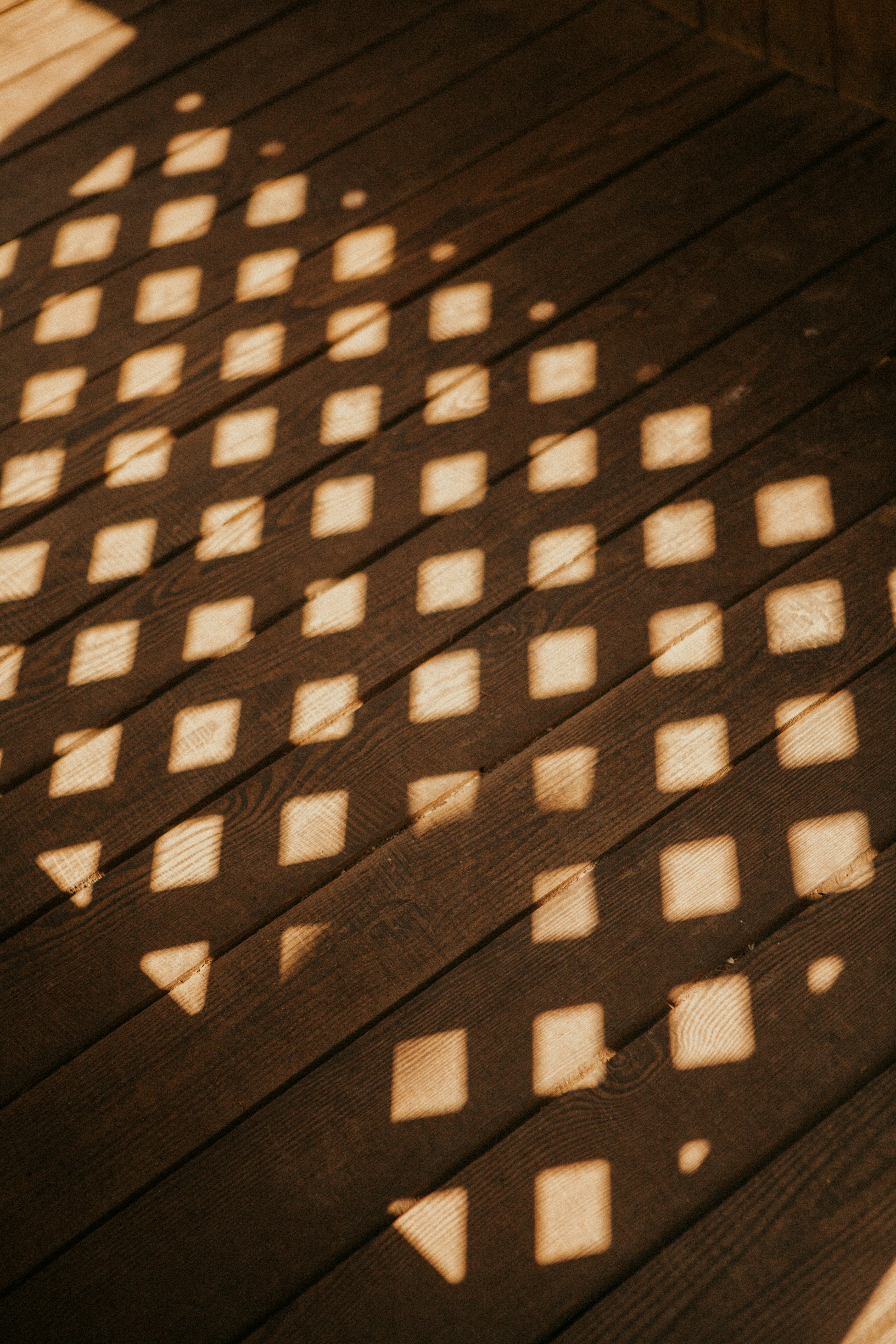 Sunlit geometric pattern of diamond gaps casts warm squares of light across dark wooden planks, creating a tactile texture and rhythmic composition.