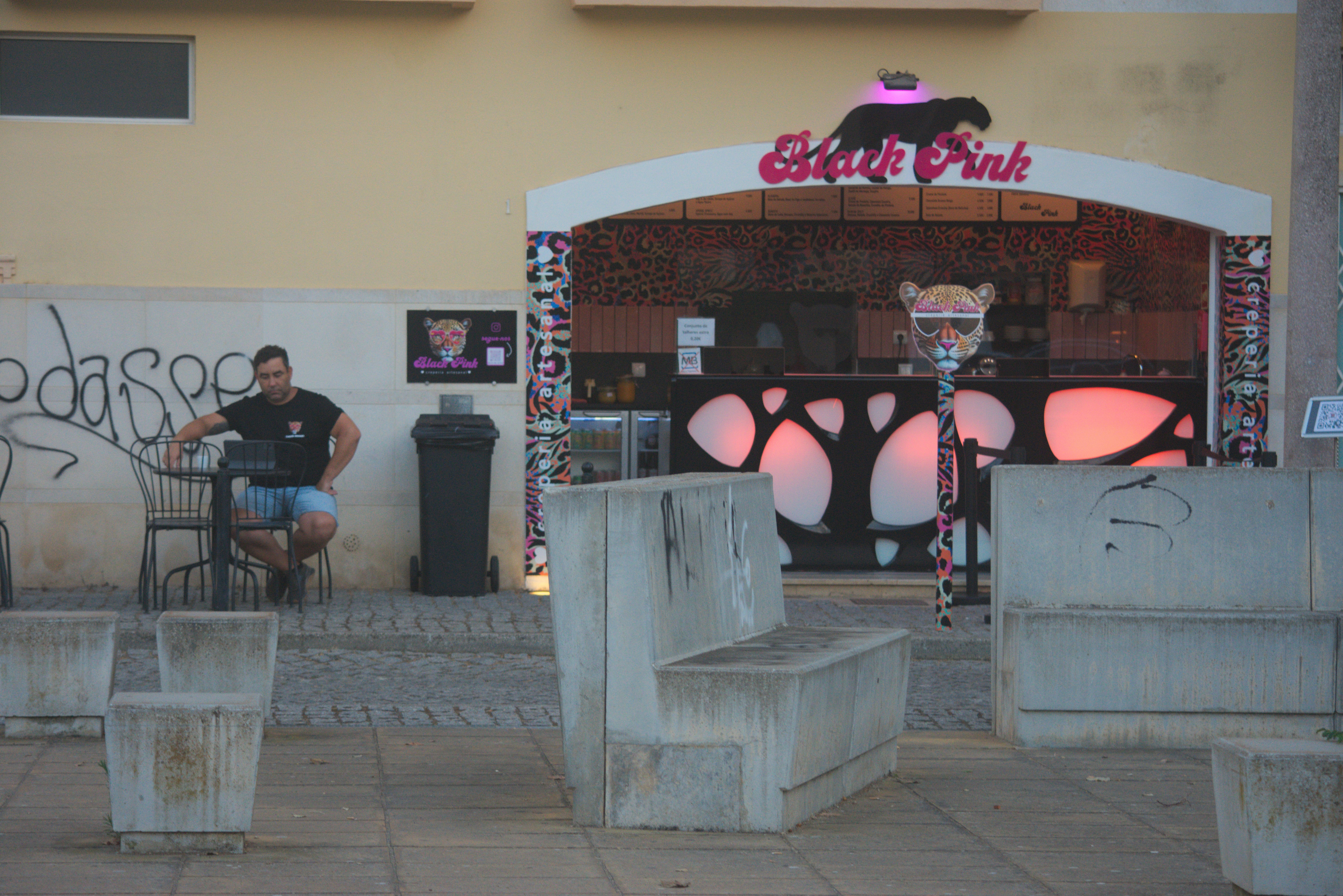 Quiet arcade storefront with a neon-lit interior and a person seated outside on the left; the scene includes concrete blocks in the foreground and a casual urban atmosphere.
