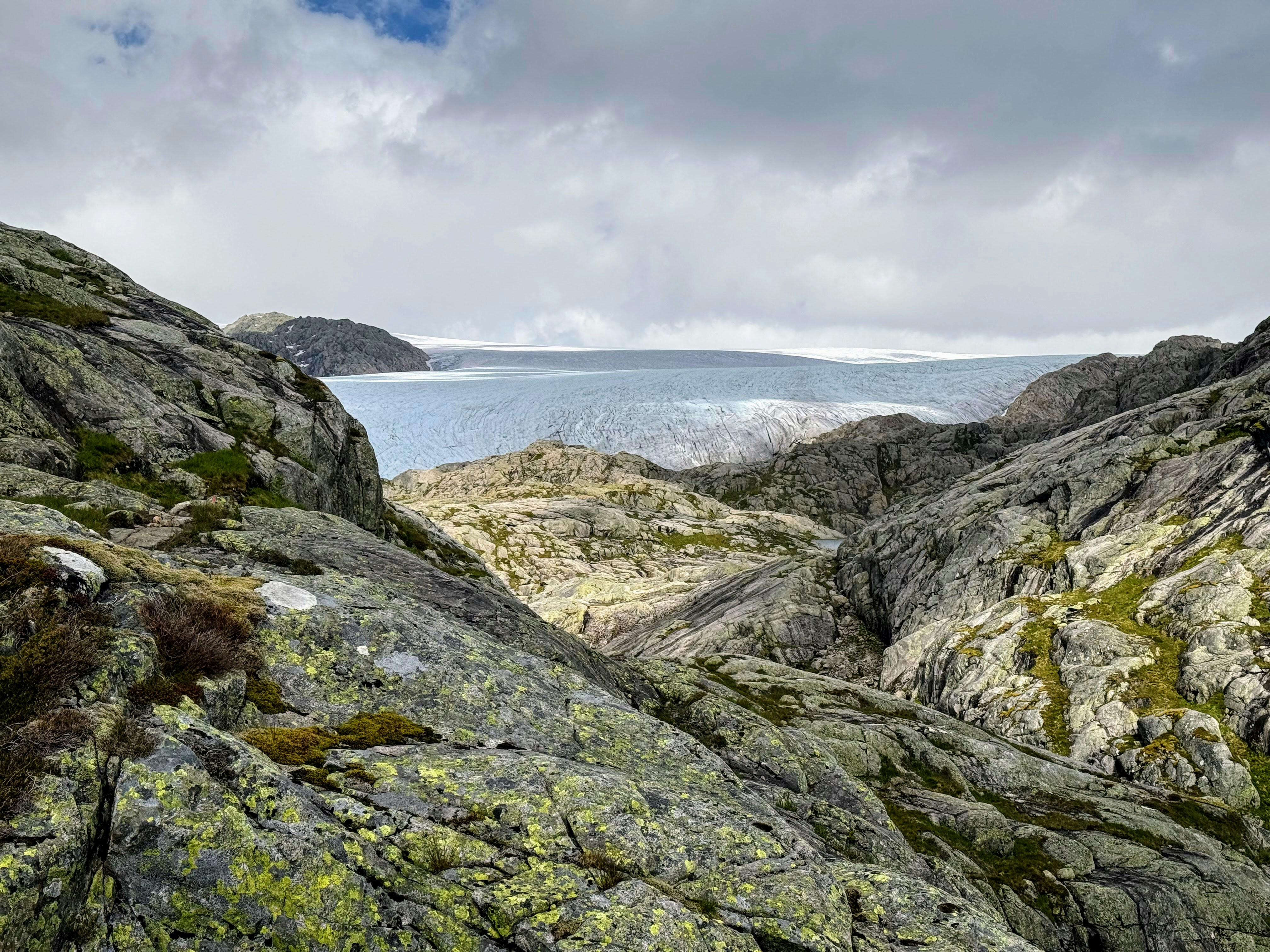 Rugged rocky shoreline with moss and lichen leads to a calm sea beyond a break in the basalt, under a cloudy sky.