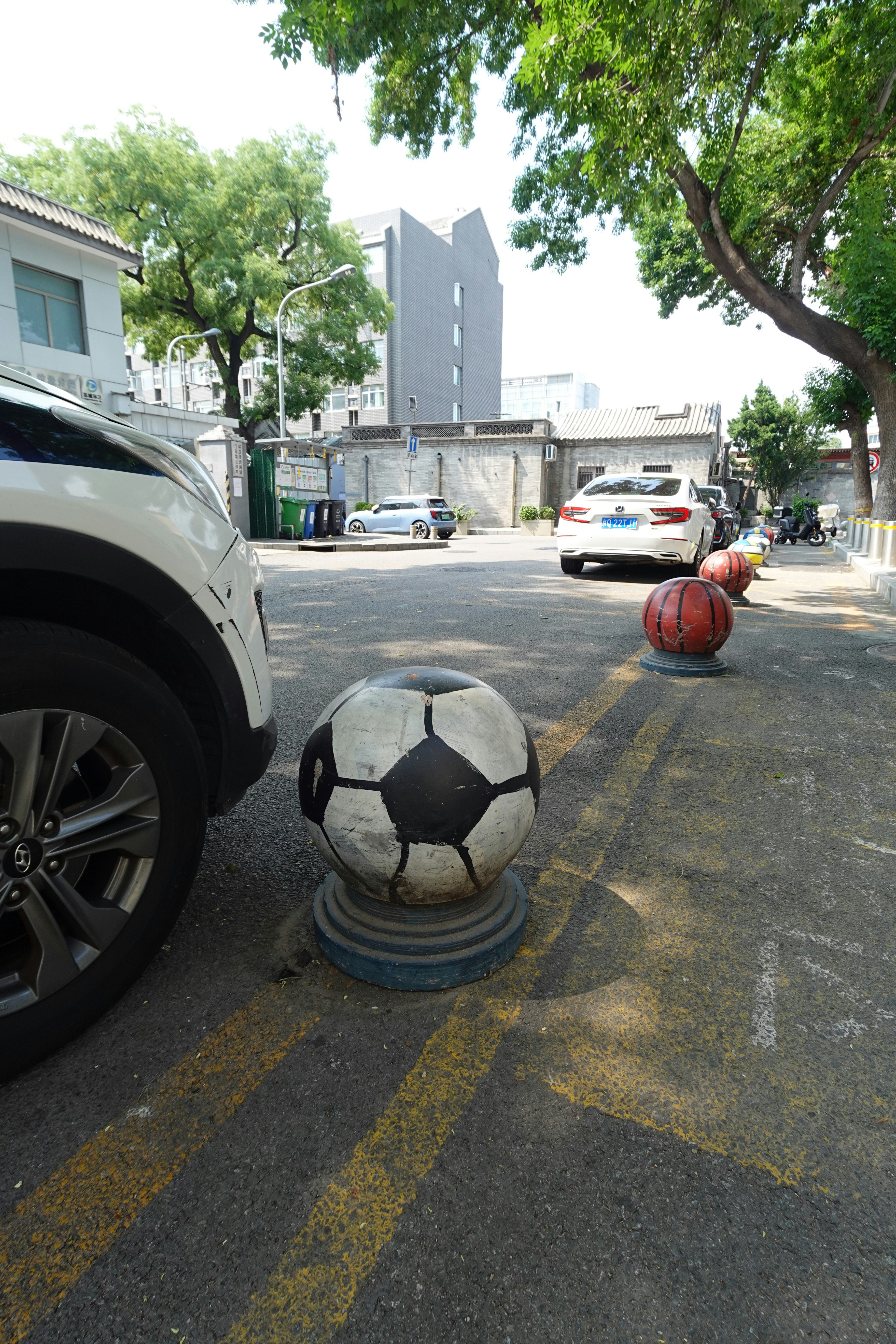Rounded soccer-ball bollard sits center foreground as cars line a sunlit street, with a white SUV on the left and a distant brick wall; a tree-framed urban scene emphasizing depth and daytime activity.