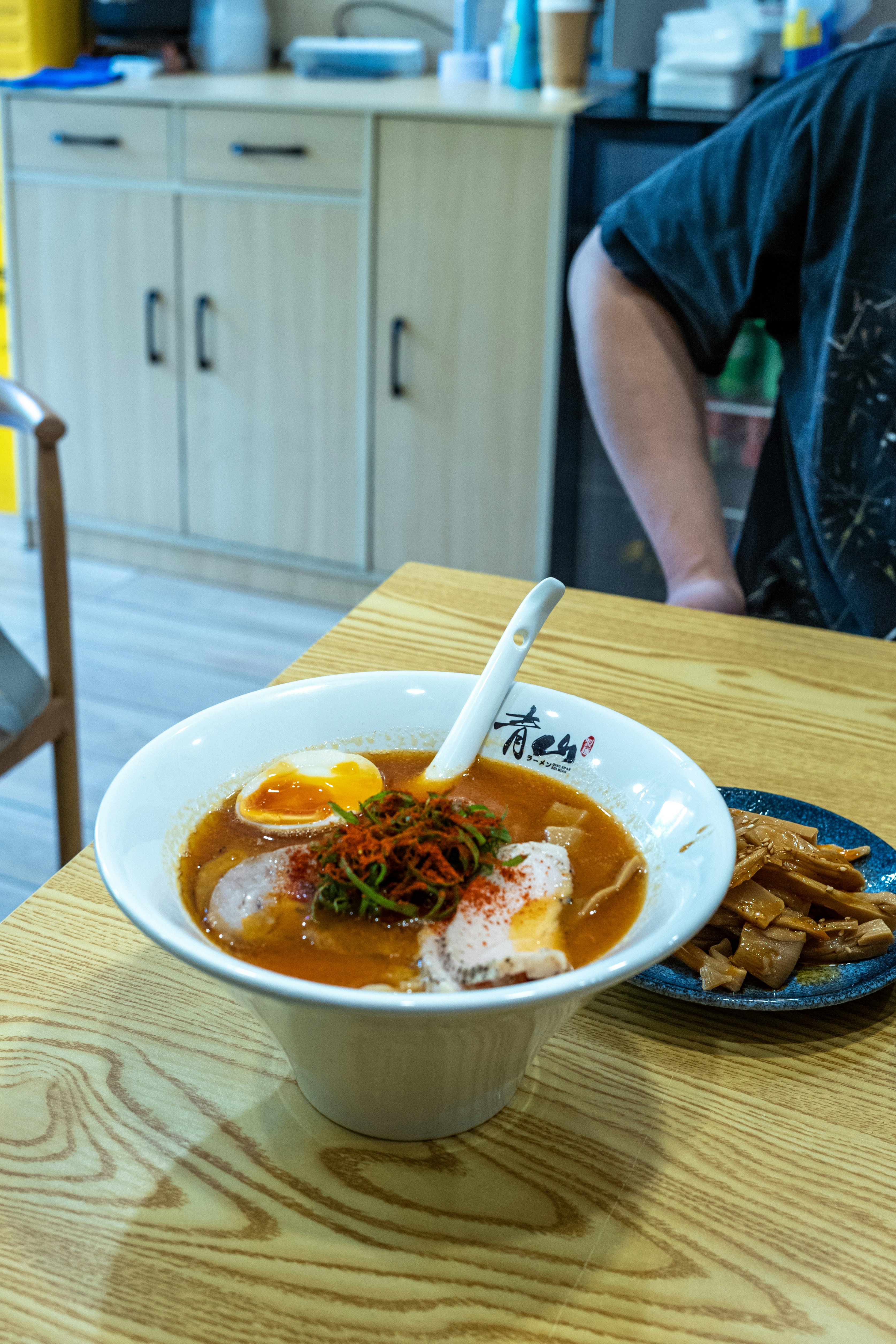 Bowl of ramen at a casual cafe with a side plate and a partial view of a person