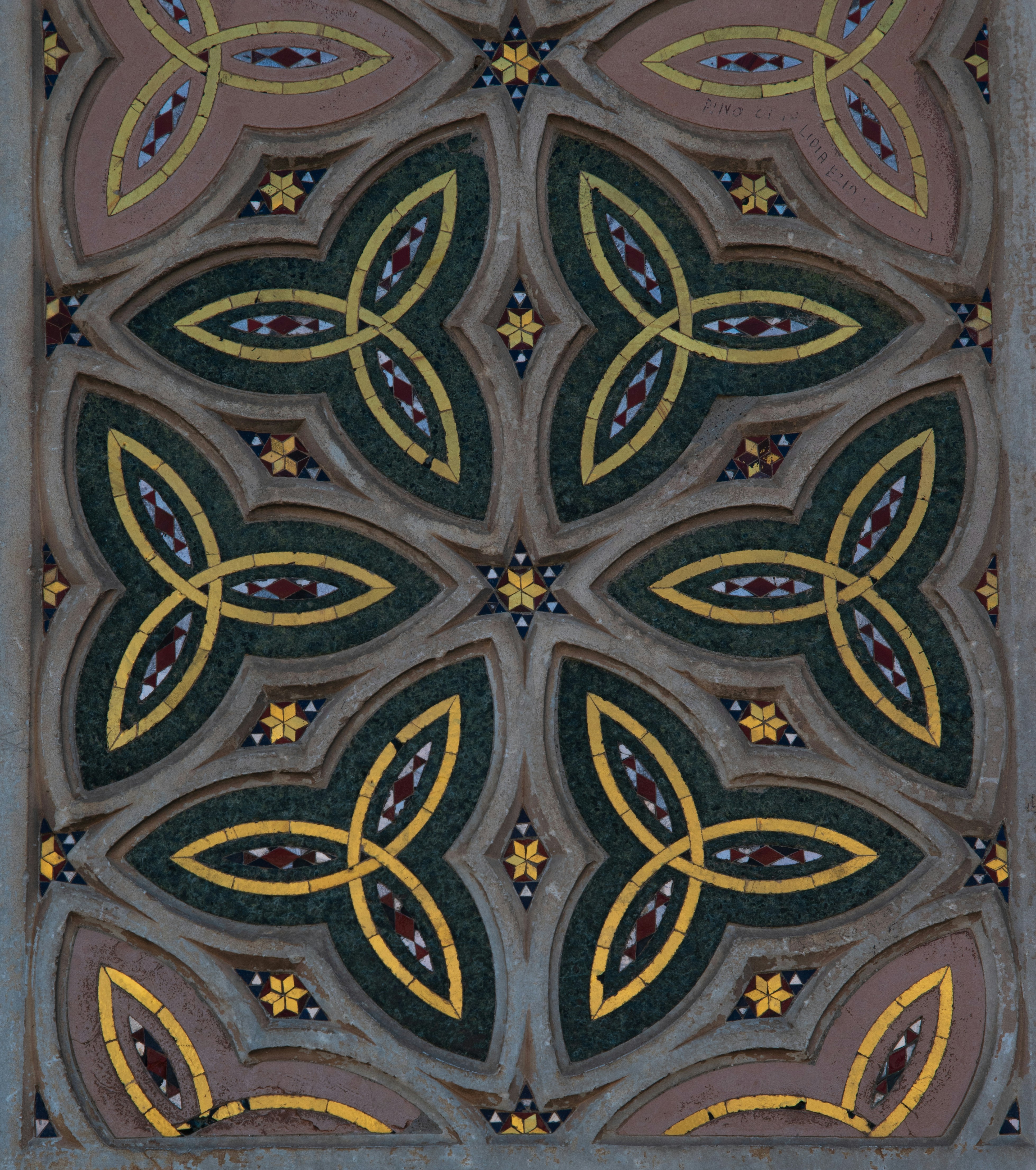 Intricate tiled motif with interlocking petal shapes in green, gold, and red, forming a symmetrical star-like pattern. Mosaic wall texture showcased as ornamental detail.