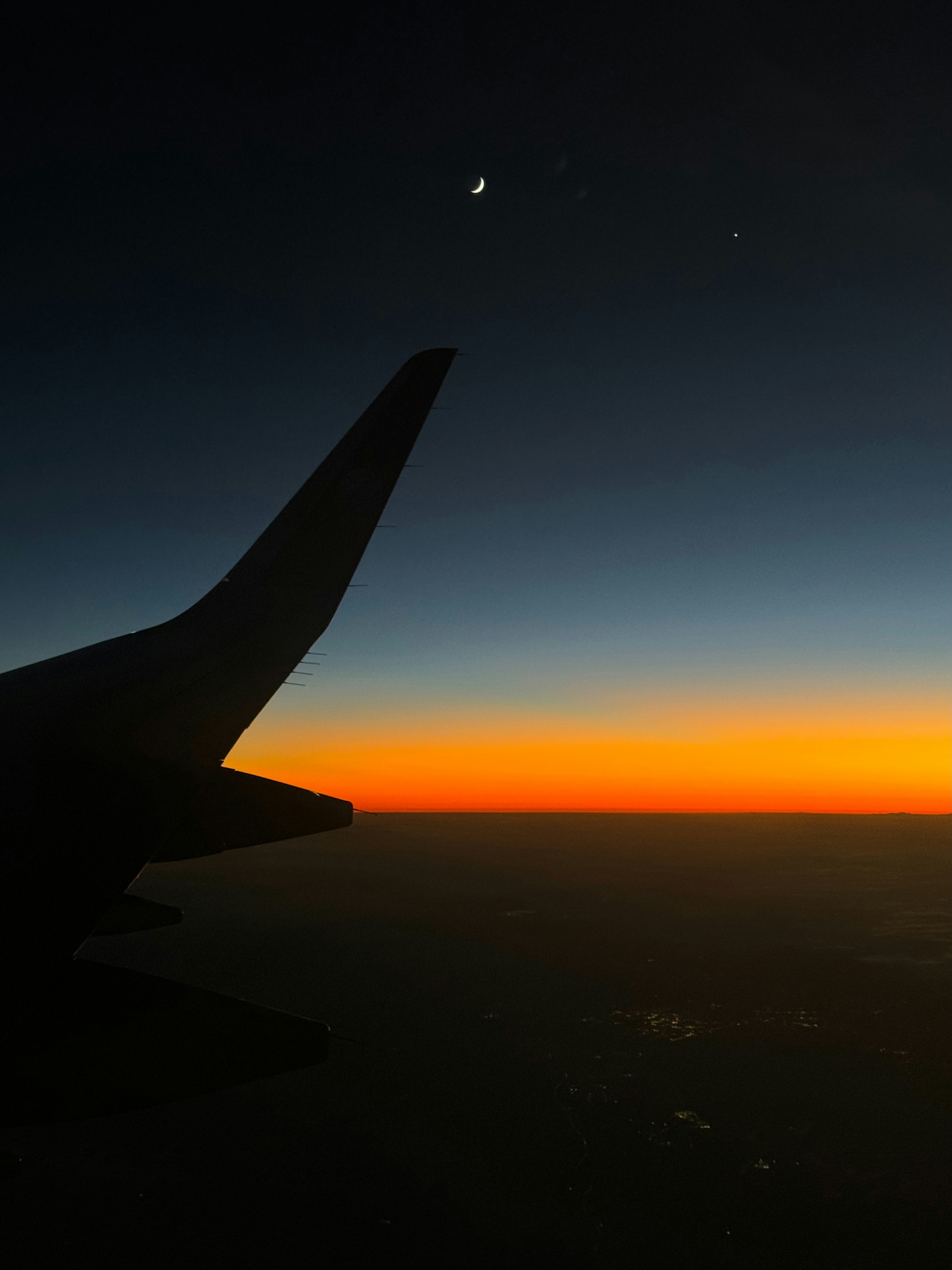 Wing silhouette against a gradient sky transitioning from deep blue to orange at sunset, with a crescent moon nearby. Aerial perspective emphasizes horizon glow and distant landscape.