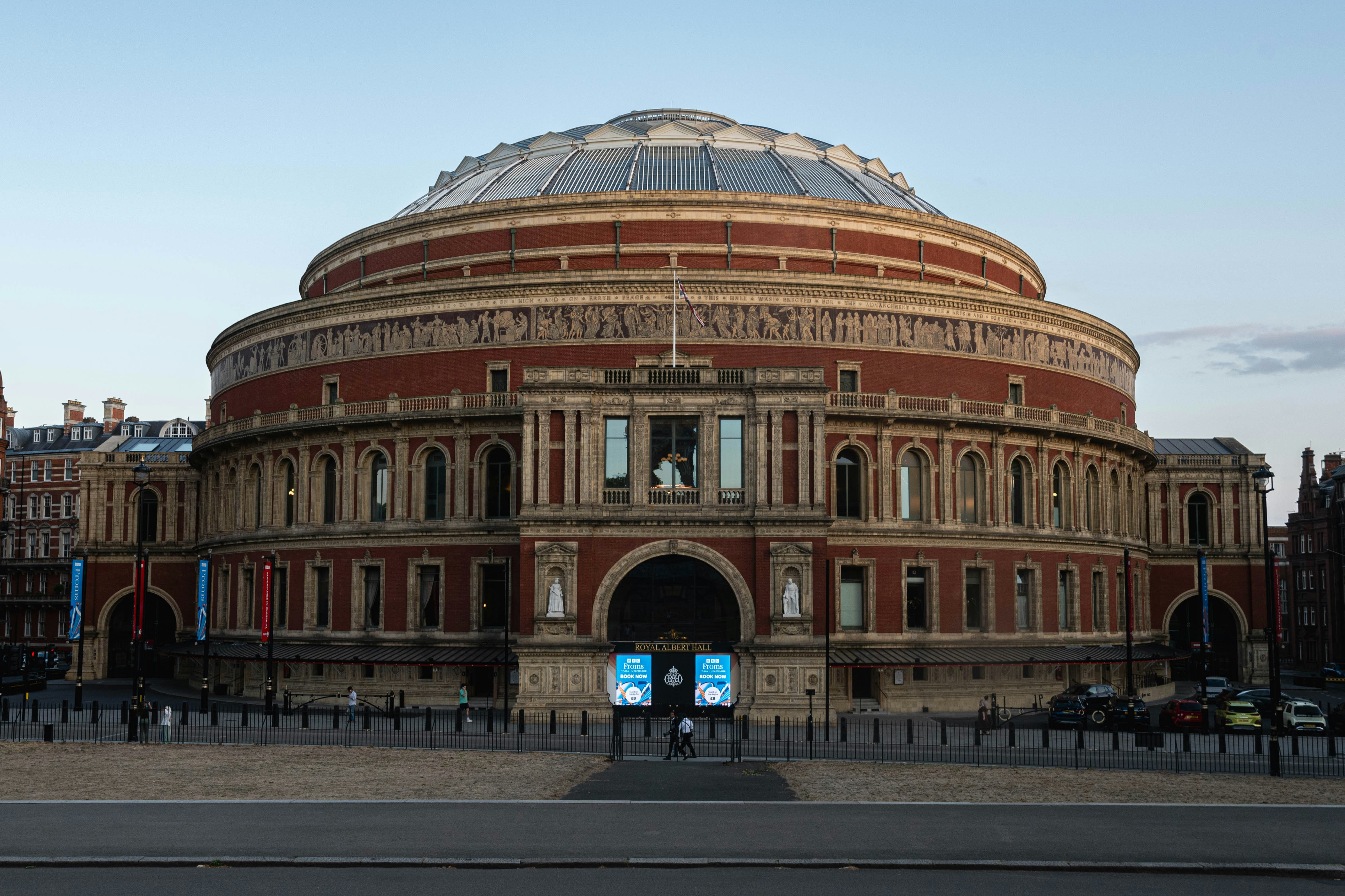 Red-brick arena with a domed roof sits centered along a riverfront promenade; two blue banners and a dark entryway mark the public-facing facade, with parked cars and pedestrians flanking the wide street. A clear sky fills the backdrop.